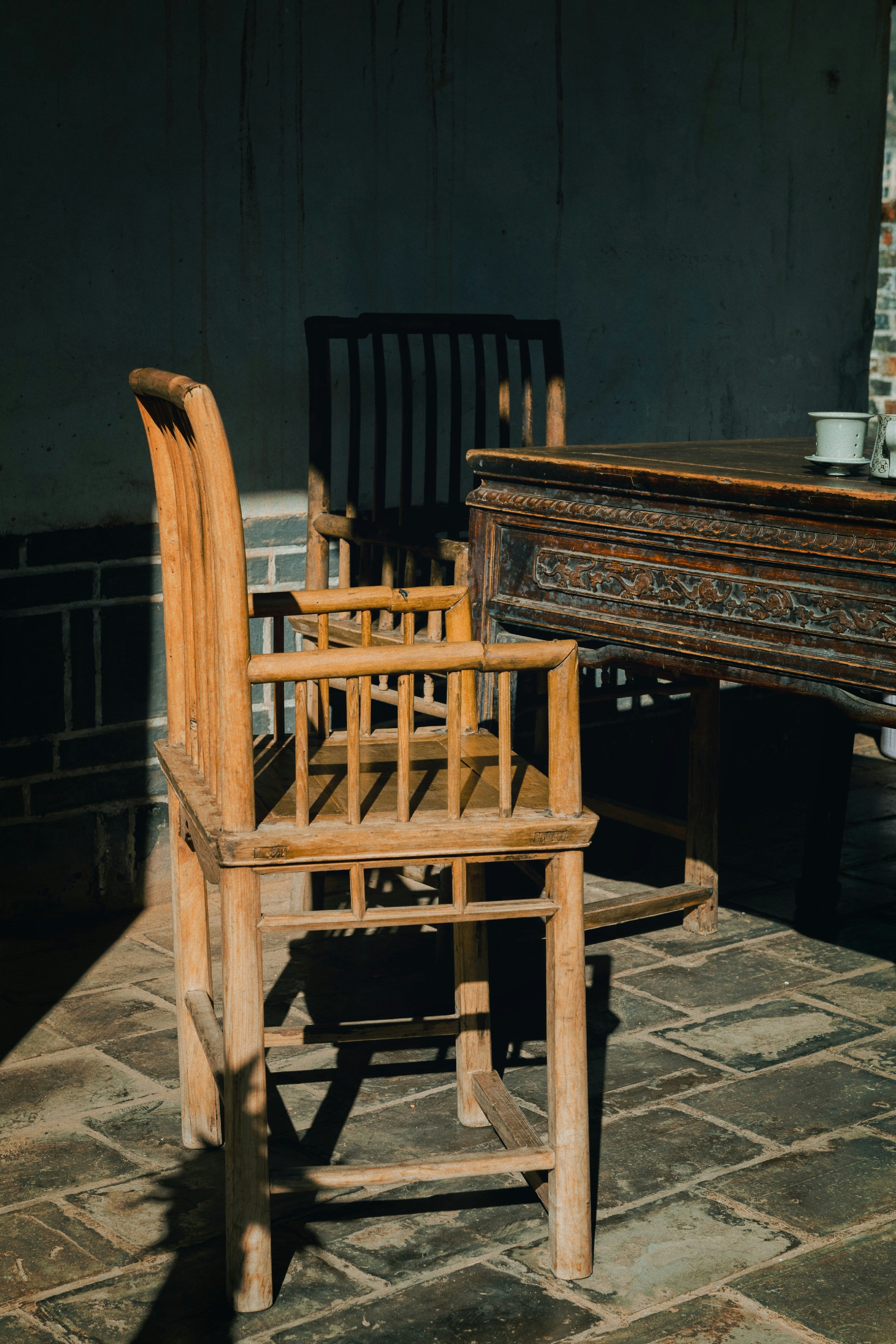 A rustic wooden chair stands beside an intricately carved table in a softly lit room, evoking a sense of history and warmth.
