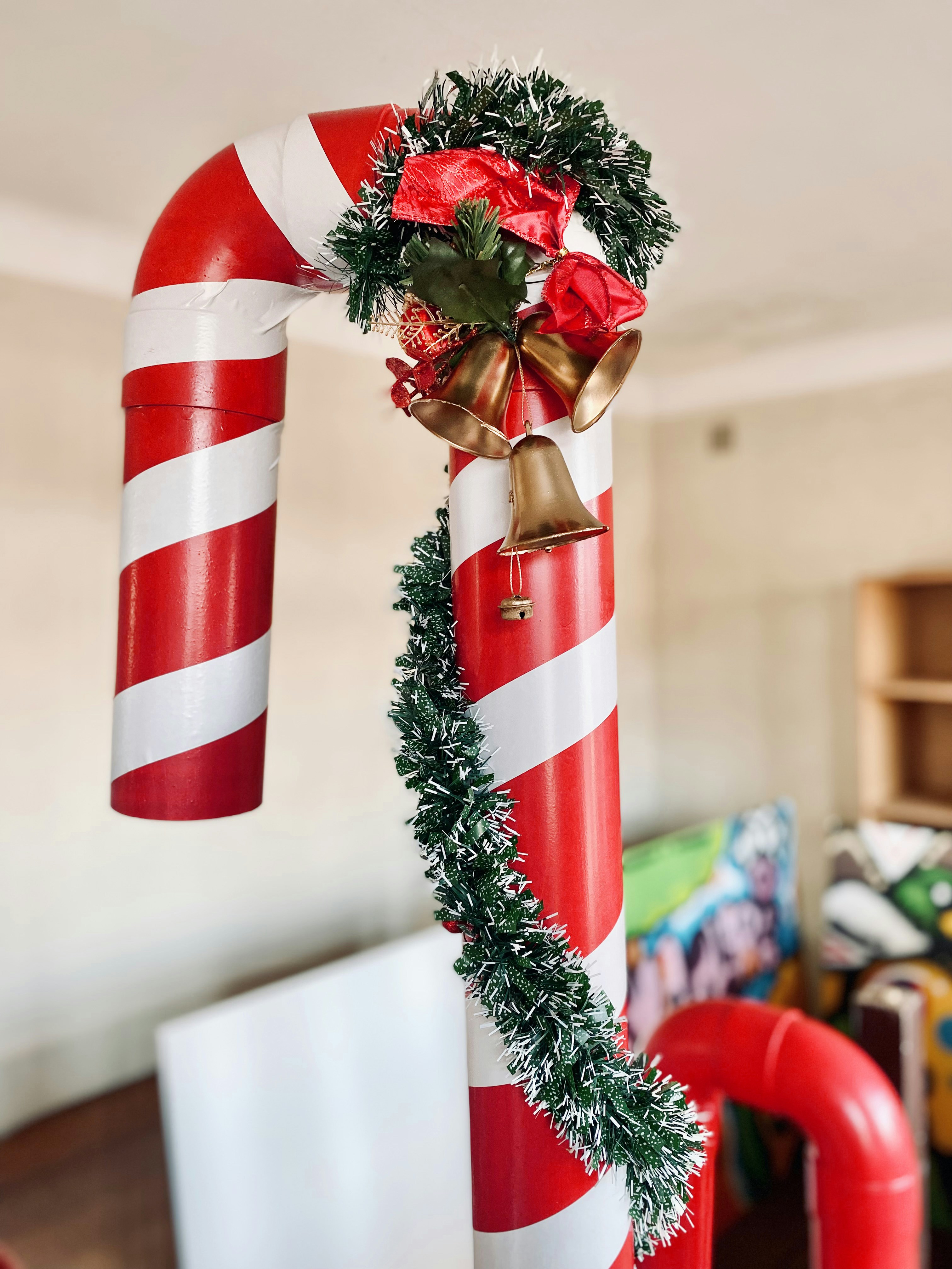 Giant candy cane decoration adorned with festive garland and bells indoors.