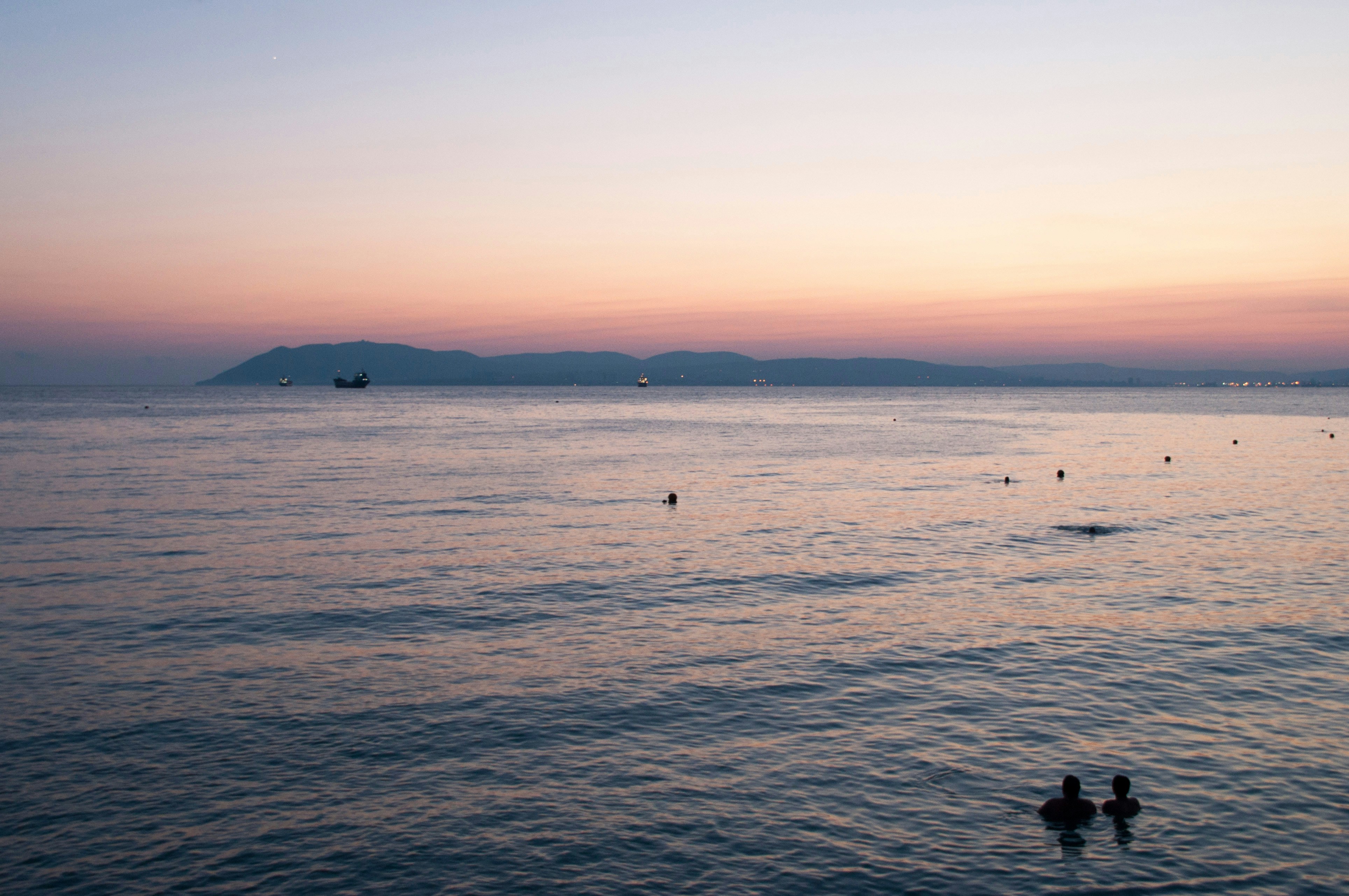 Two silhouettes of bathers enjoying the calm sea at twilight, with distant mountains and a soft gradient sky. 