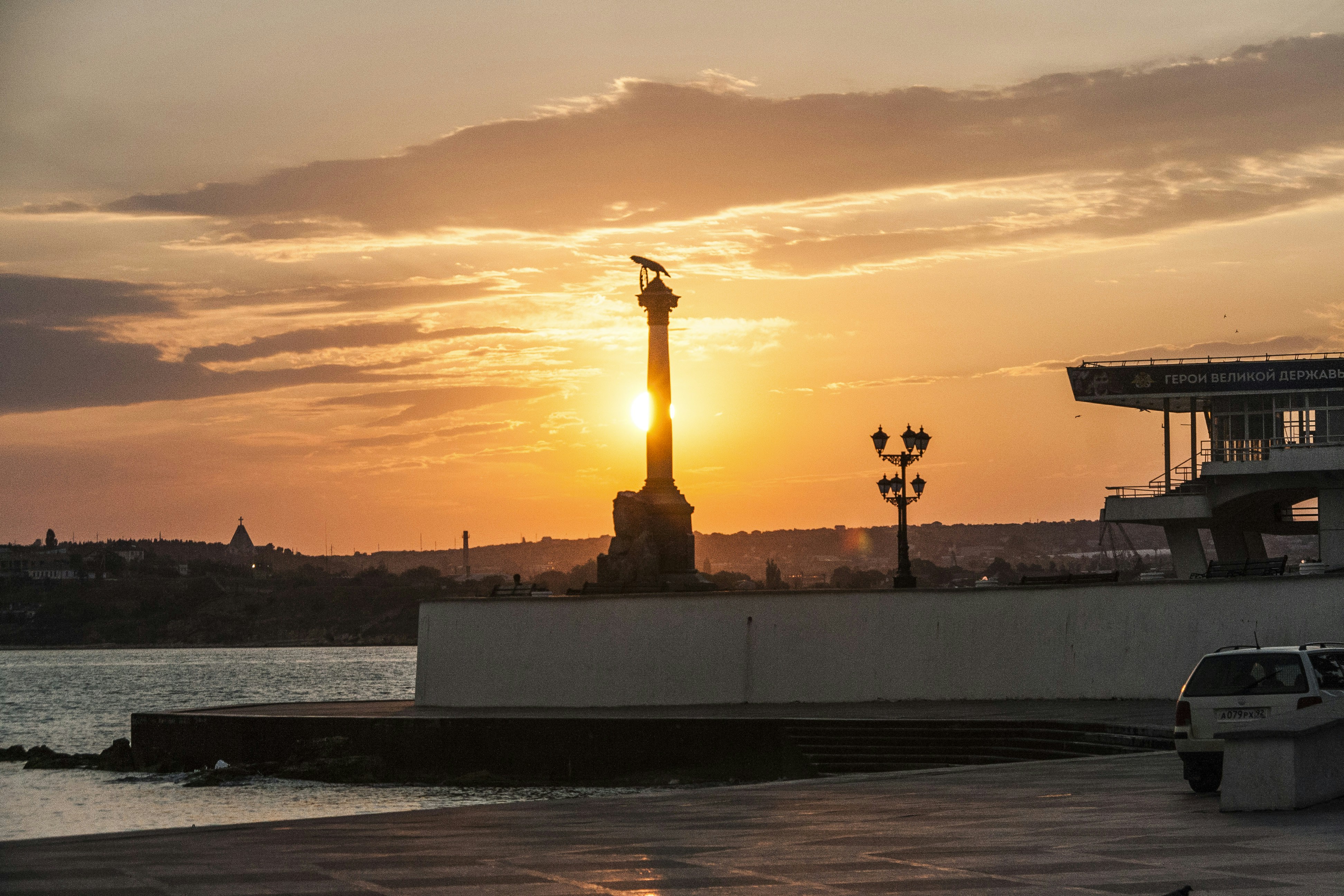 A monument silhouetted against a vibrant sunset, casting reflections on the water's surface. The scene captures the tranquil beauty of dusk by the coast.