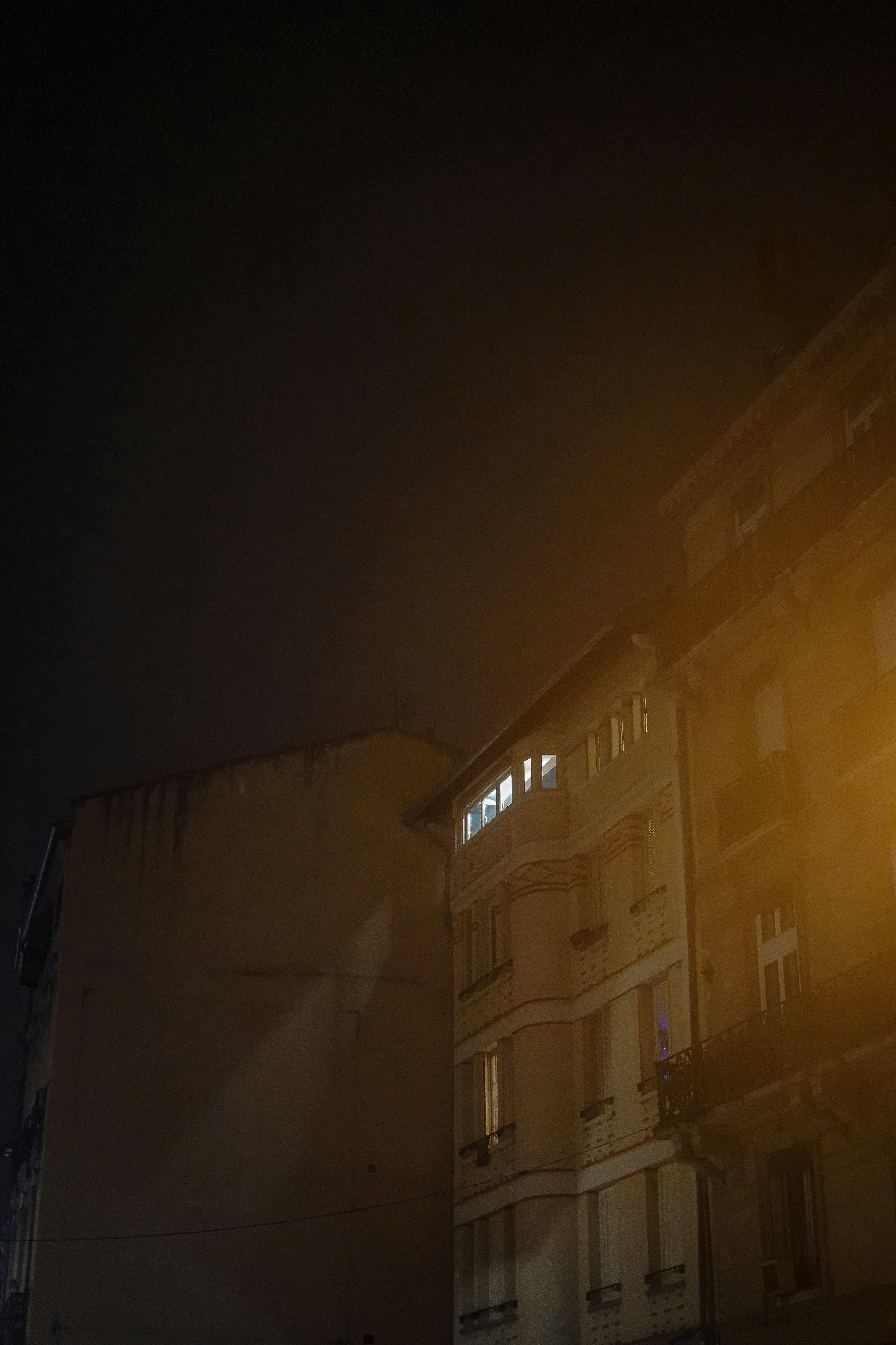 Dimly lit urban scene featuring a building facade partially obscured by fog, with a single window glowing in the darkness.