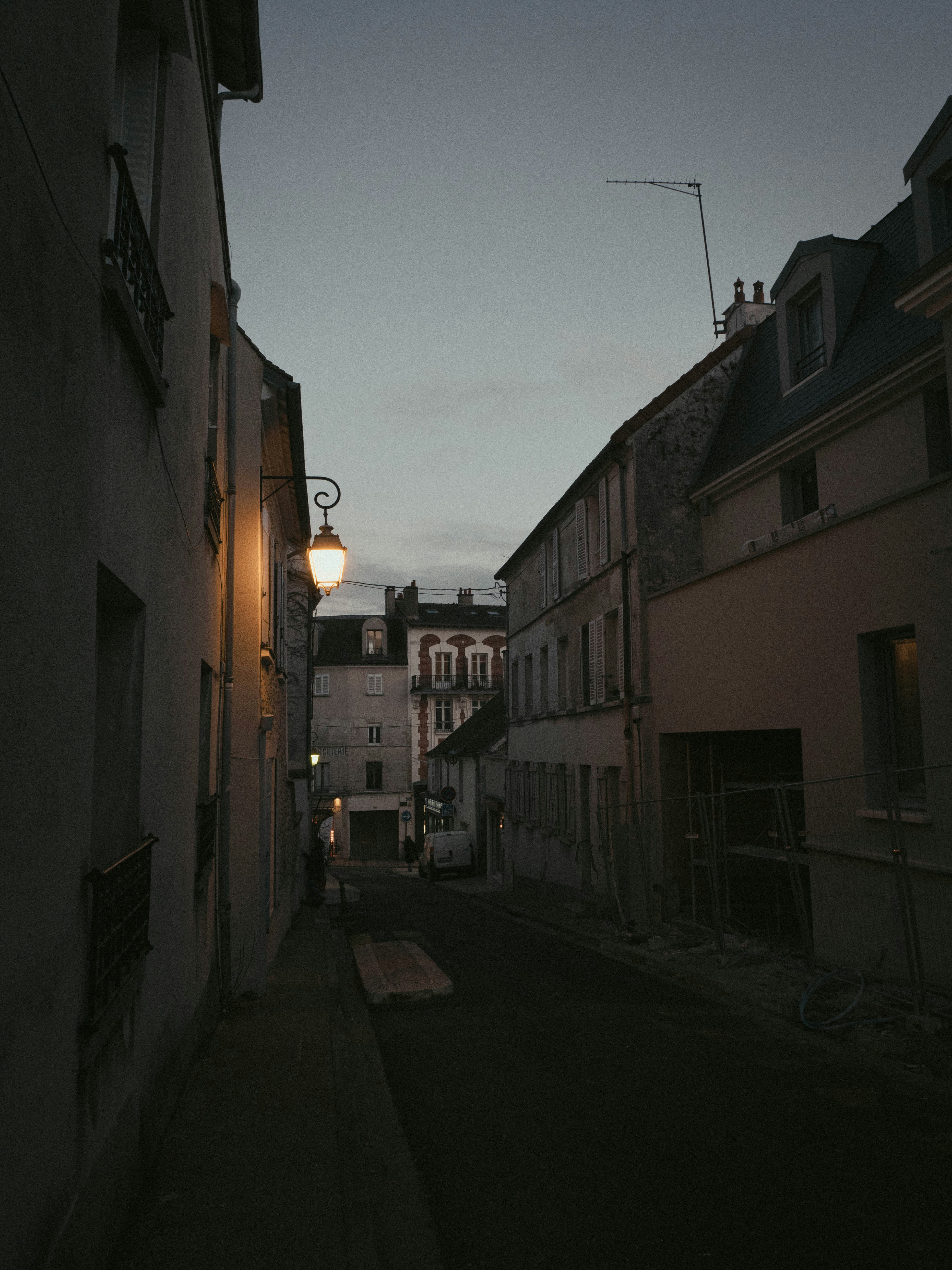 Narrow cobblestone alley illuminated by a vintage street lamp, leading to distant buildings under a twilight sky.