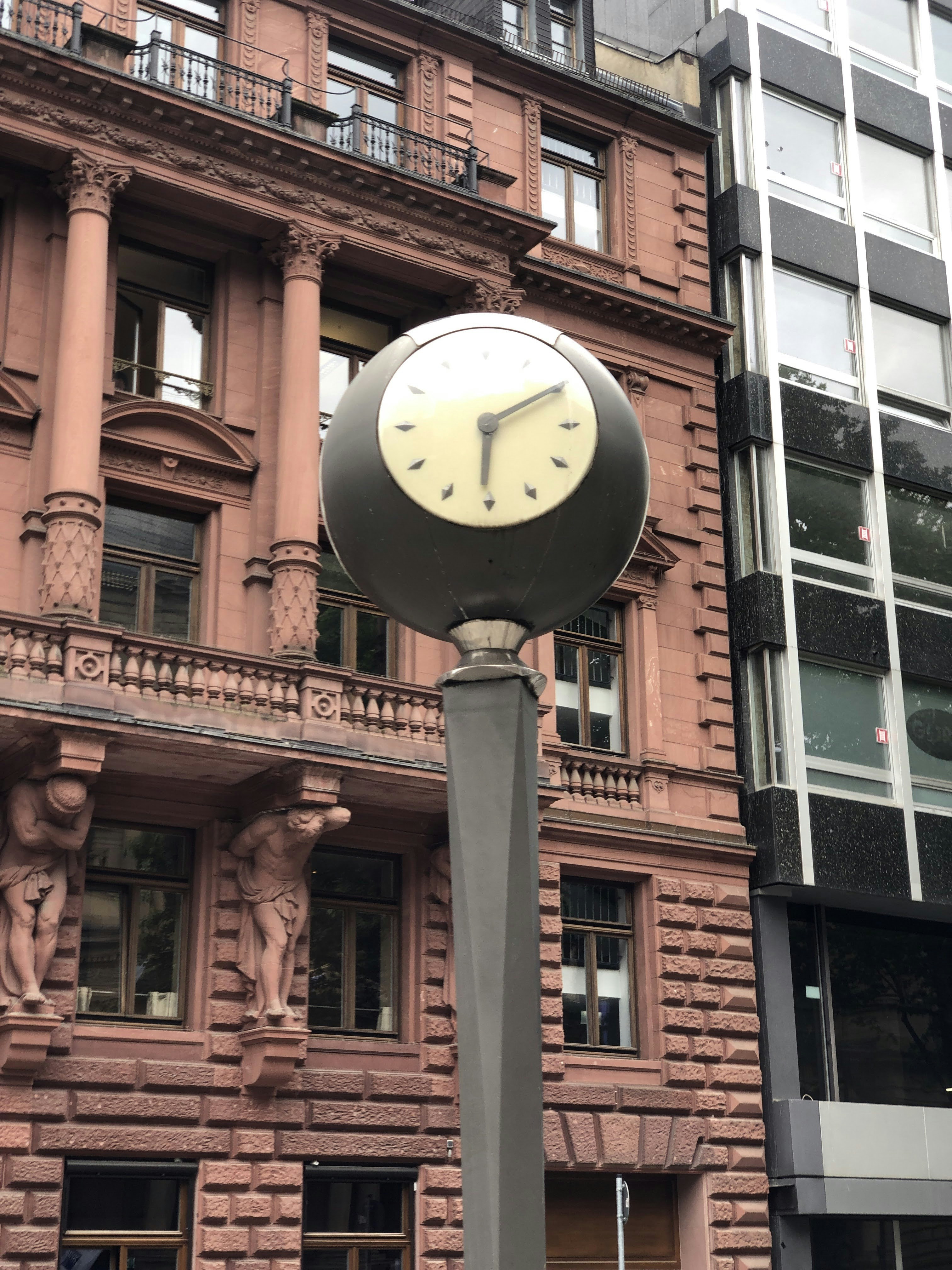 A contemporary clock mounted on a sleek pole in front of an ornate historical building, showcasing a blend of modern and classical design elements.