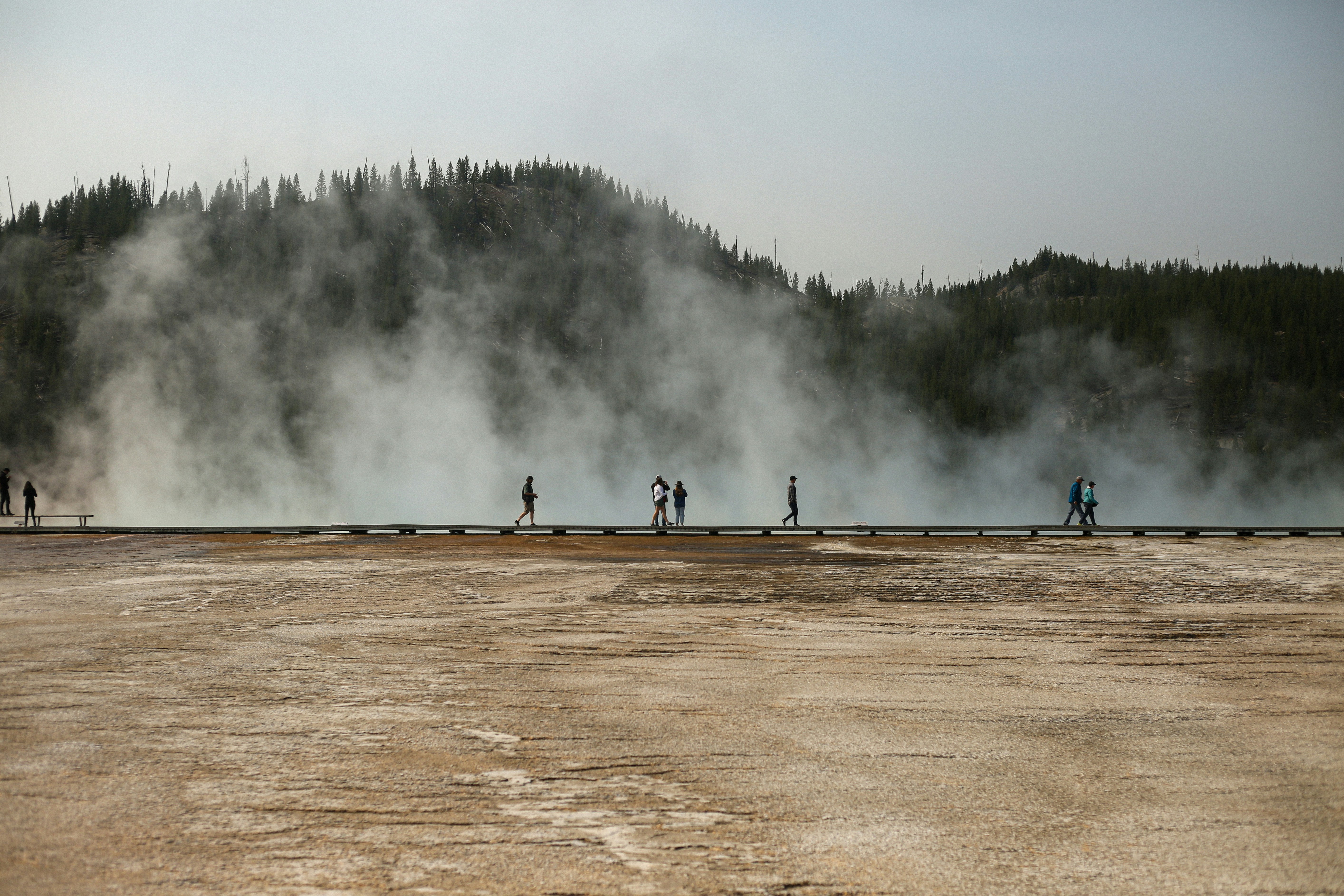 Visitors stroll along the boardwalk near a thermal spring, enveloped in steam with a backdrop of lush greenery.
