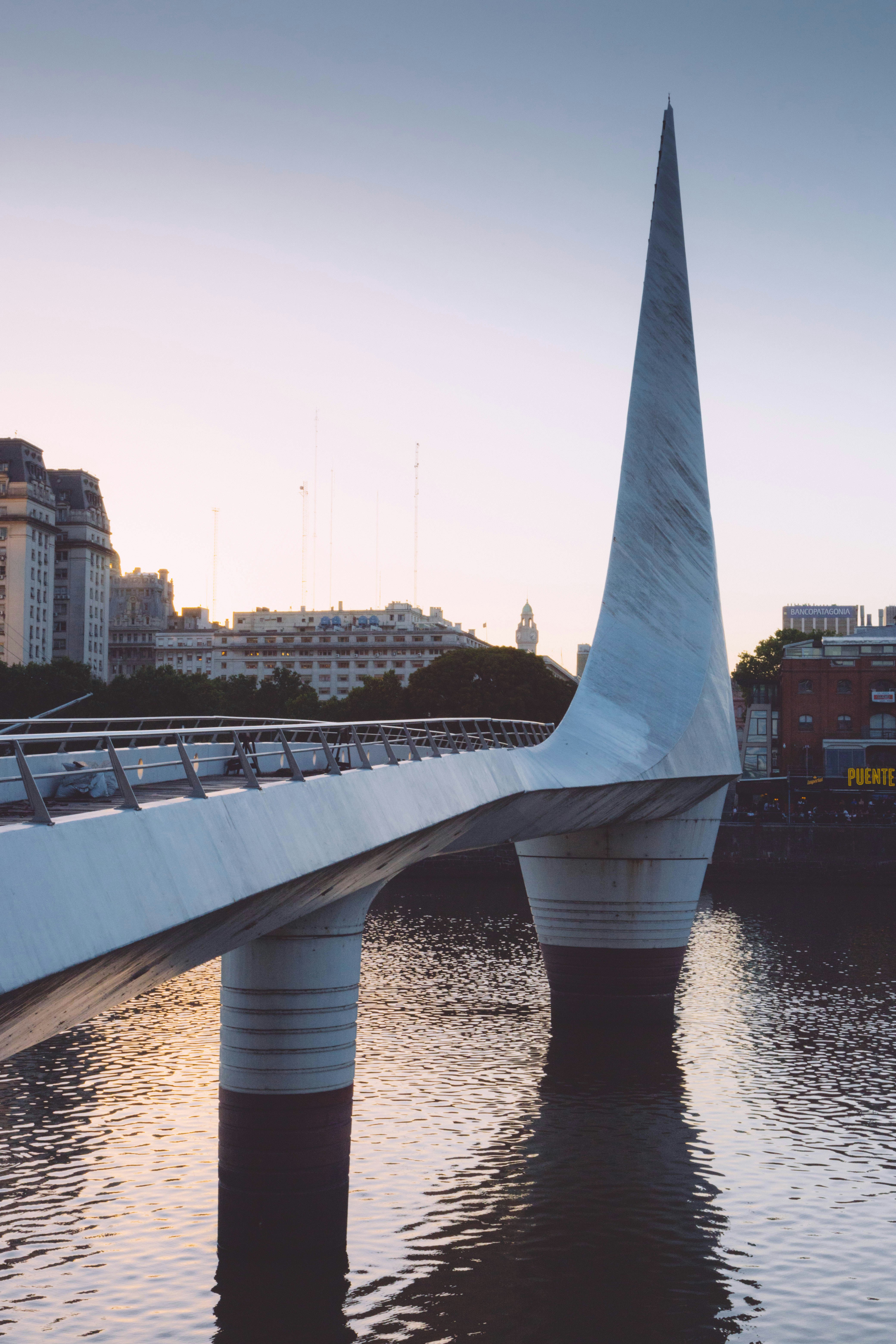 A modern bridge spans a tranquil river, showcasing its sleek design against a pastel sky. The reflection in the water enhances its architectural beauty.