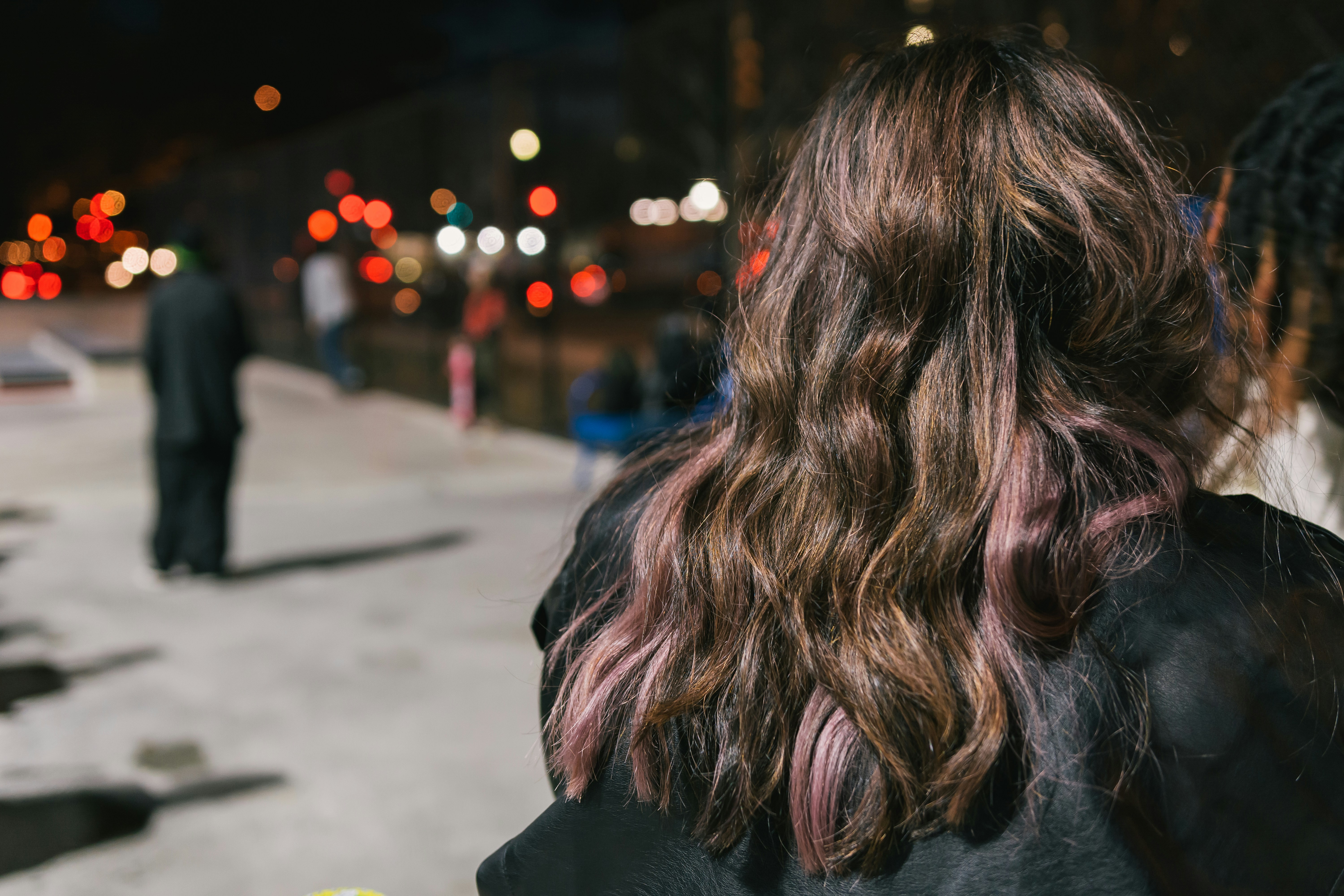 Woman with wavy hair stands with her back to the camera, surrounded by a vibrant city nightlife backdrop. The blurred lights of vehicles and pedestrians create a dynamic atmosphere.