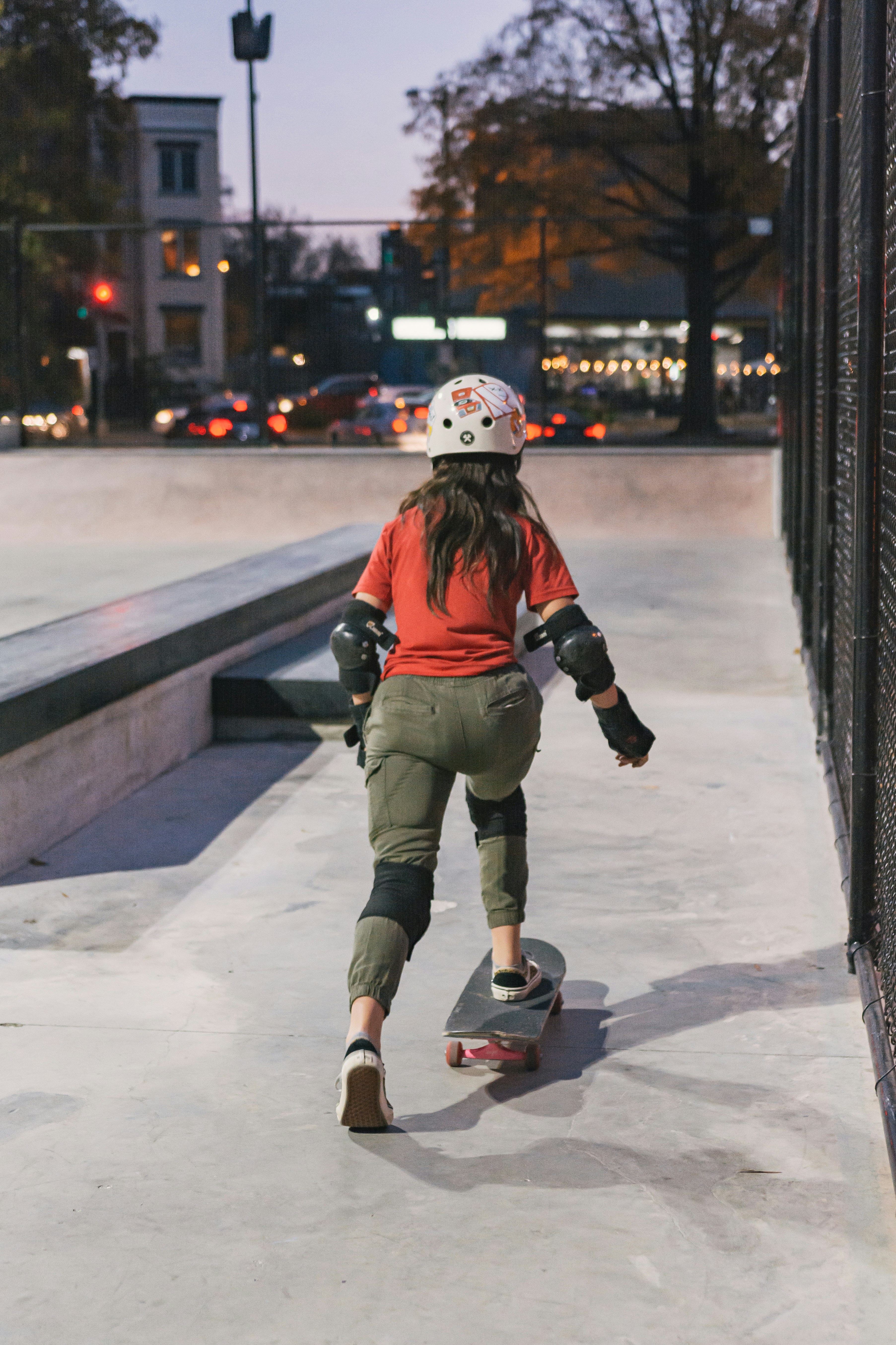Young skateboarder in protective gear gliding through a skatepark at dusk, with city lights glowing in the background.