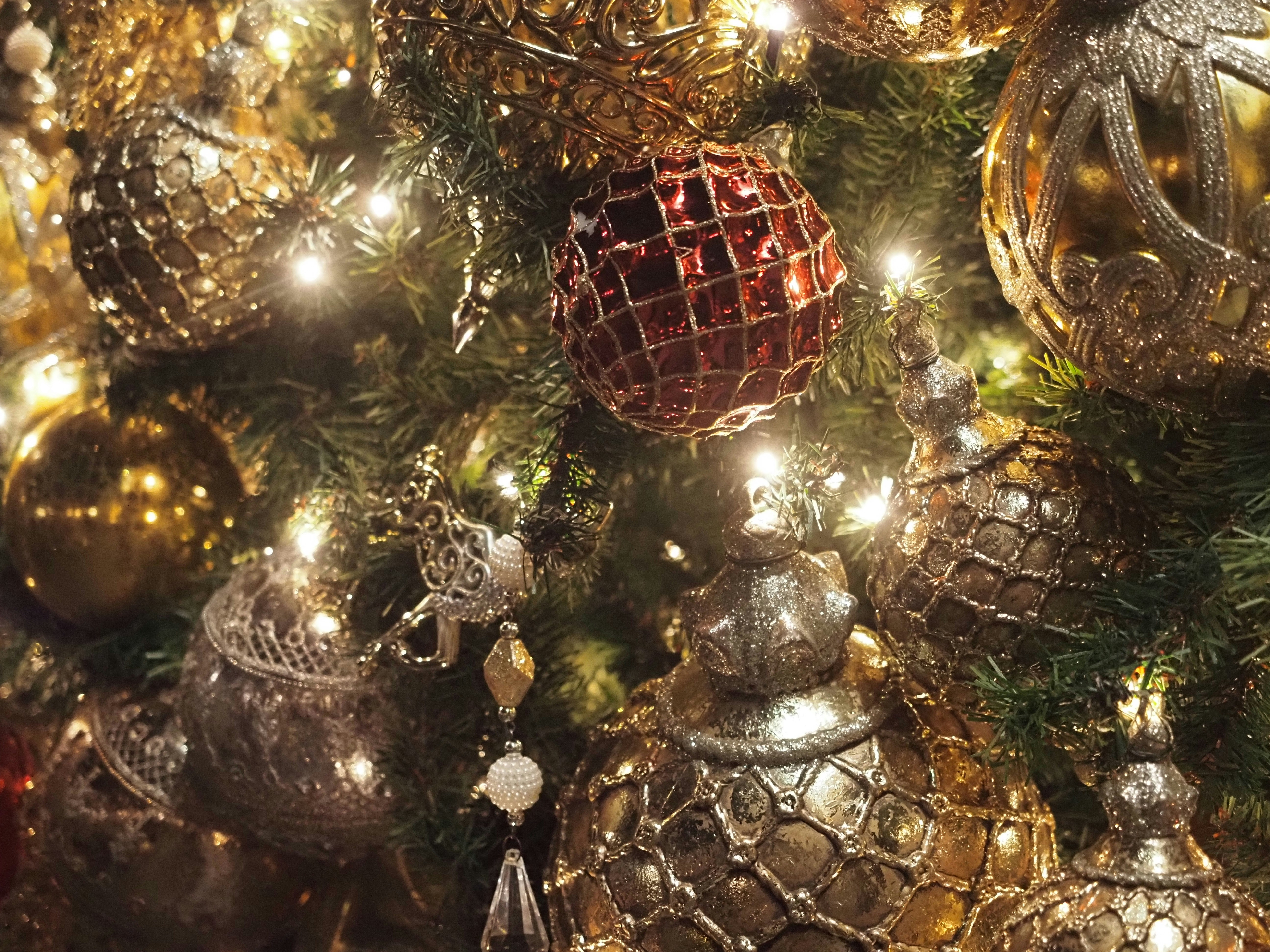 Close-up photograph of metallic Christmas baubles and warm fairy lights on a pine tree, with a red mosaic orb at center.