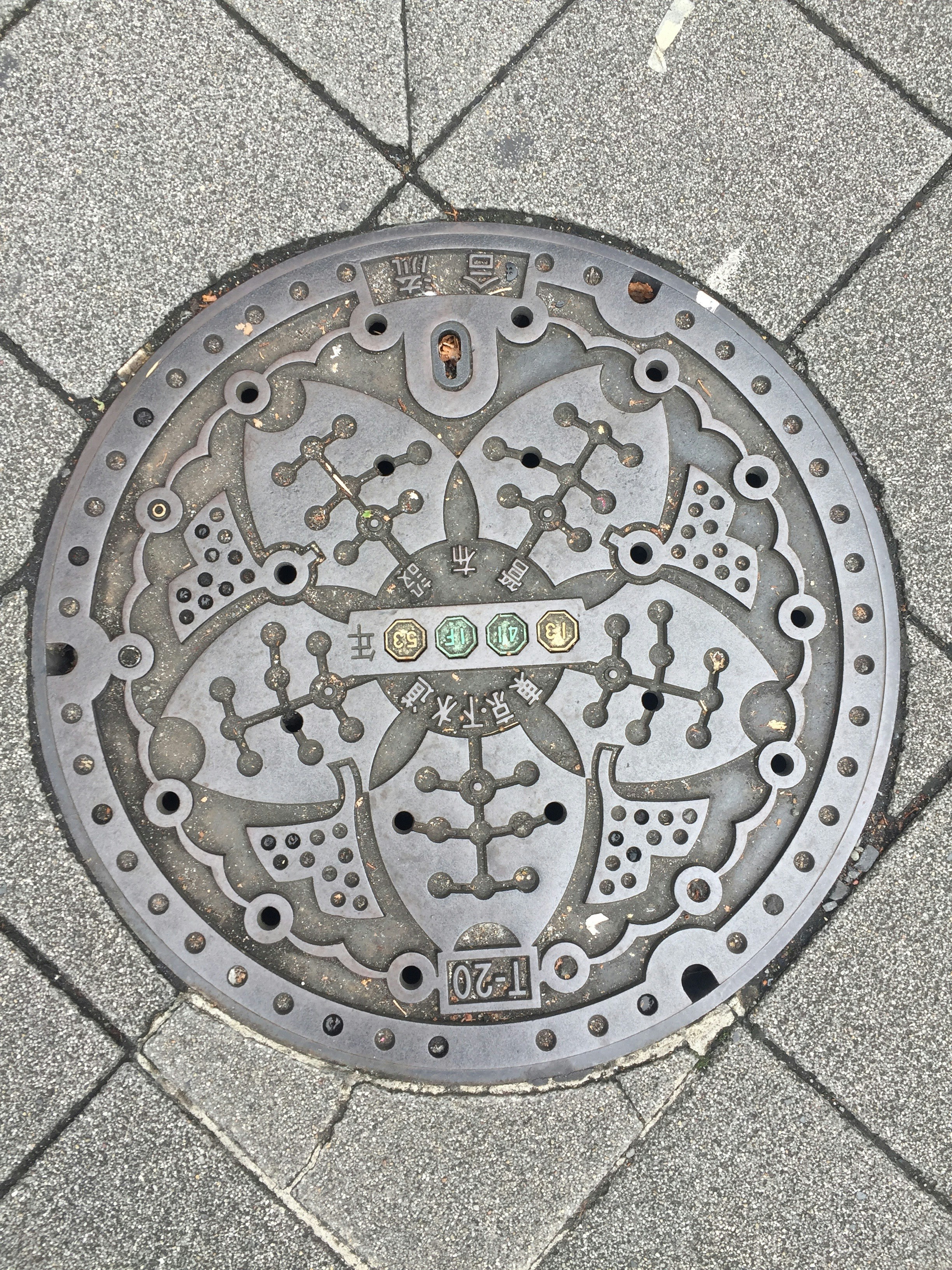 Intricate manhole cover featuring a decorative design and symbols, set against a textured pavement background.