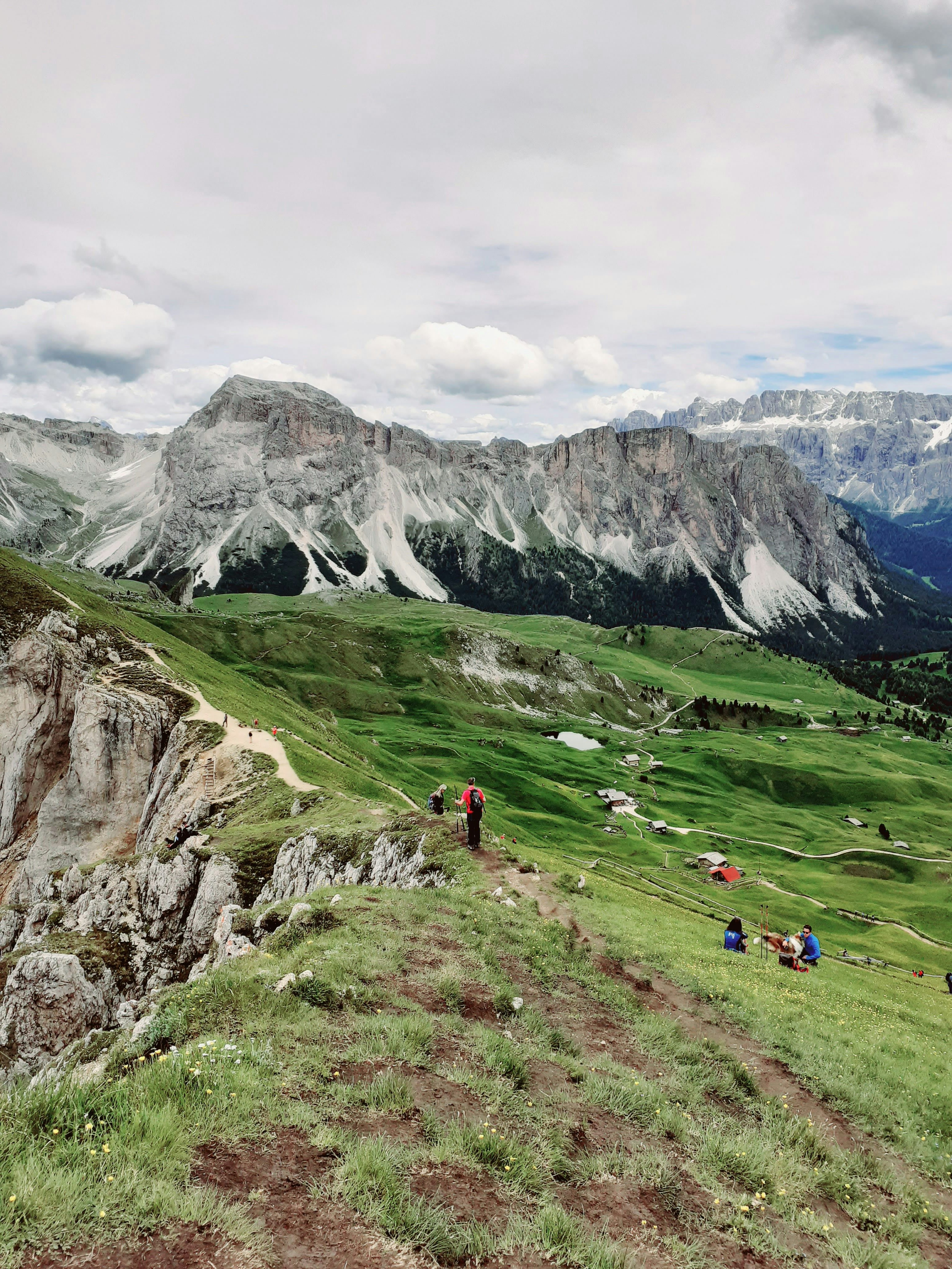 Hikers traverse a grassy alpine trail with rugged, snow-dusted peaks looming in the distance.