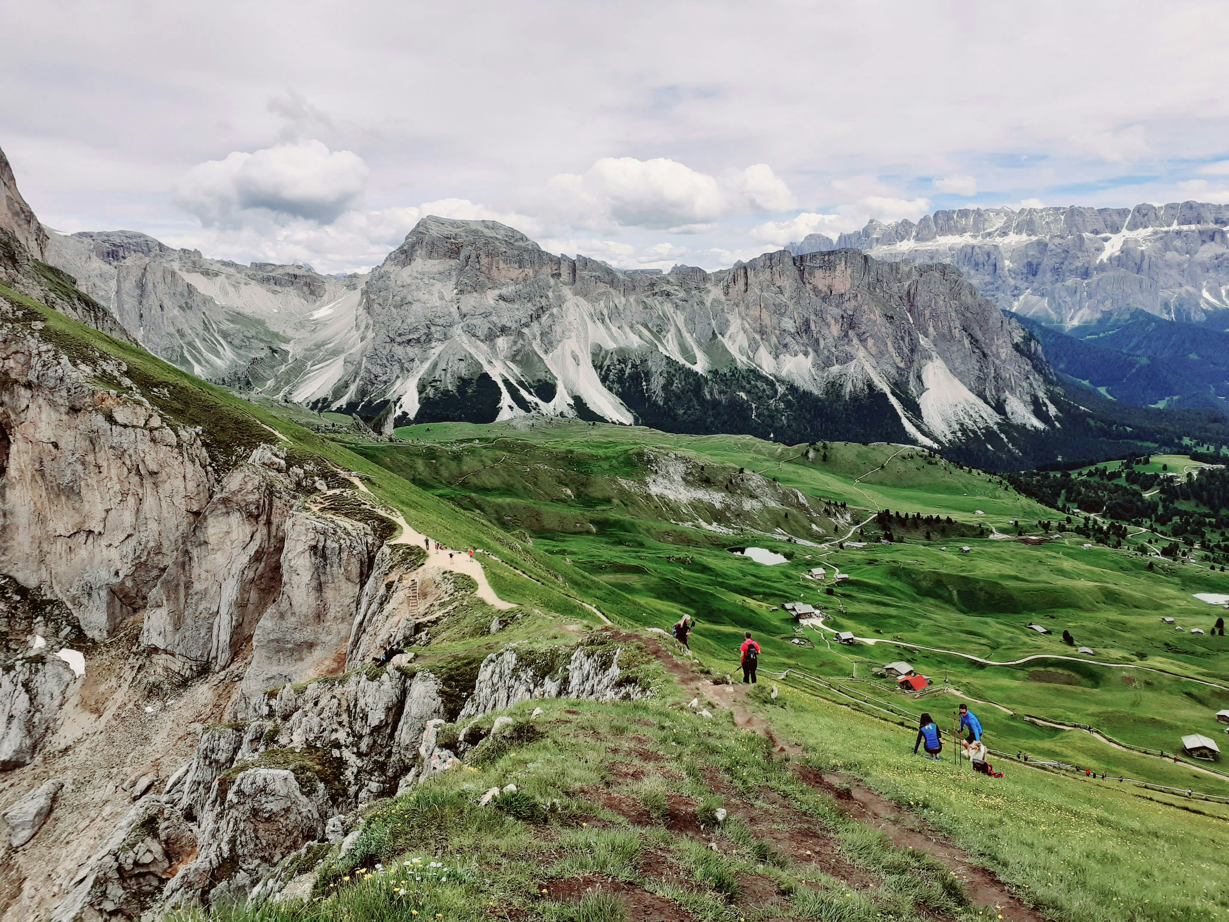Hikers traverse a lush green trail with rugged mountains in the background under a cloudy sky.