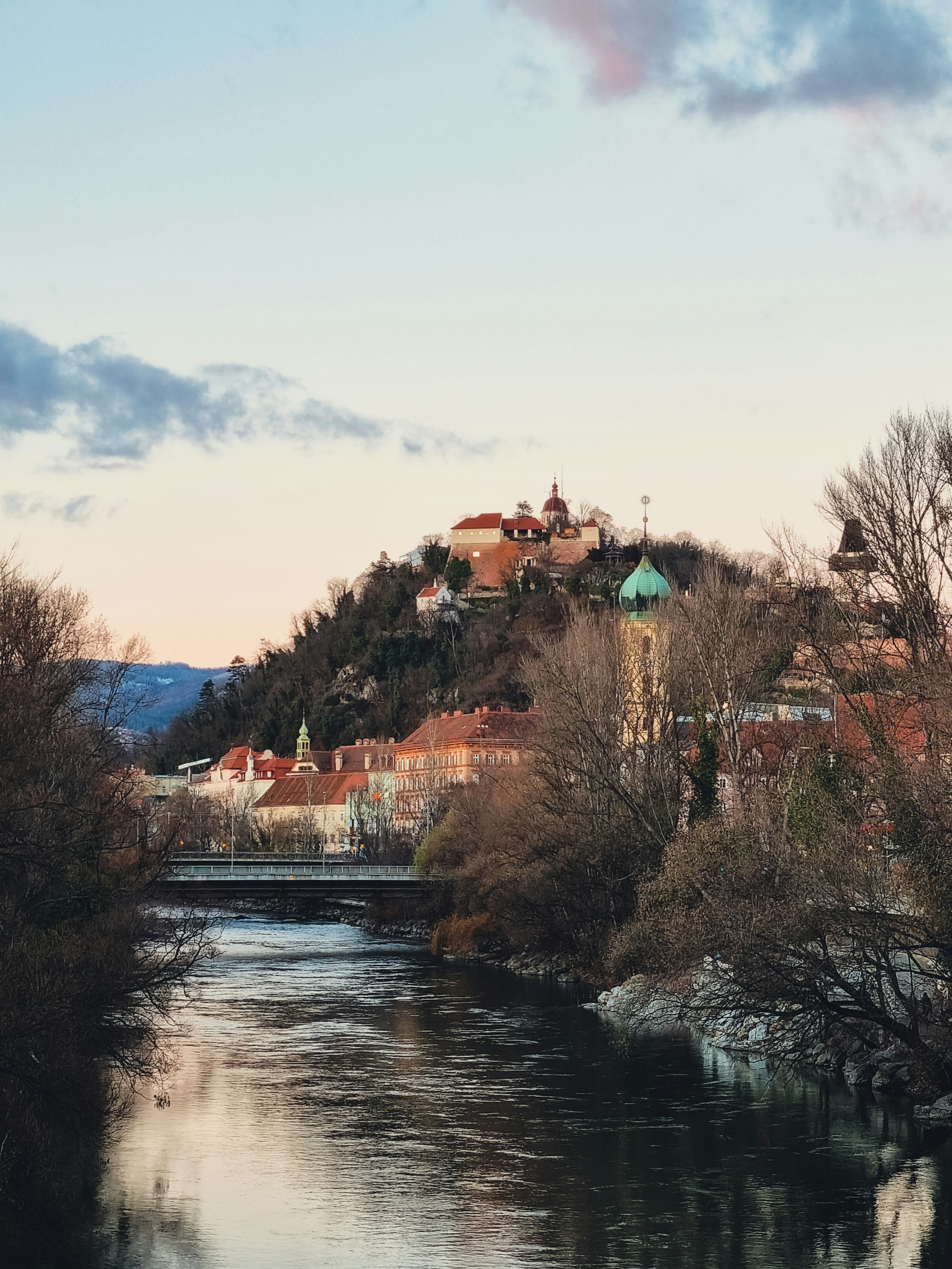 Historic hilltop castle overlooking a serene river, framed by autumn trees and a pastel sky. The scene captures the tranquility of nature and architecture harmoniously.