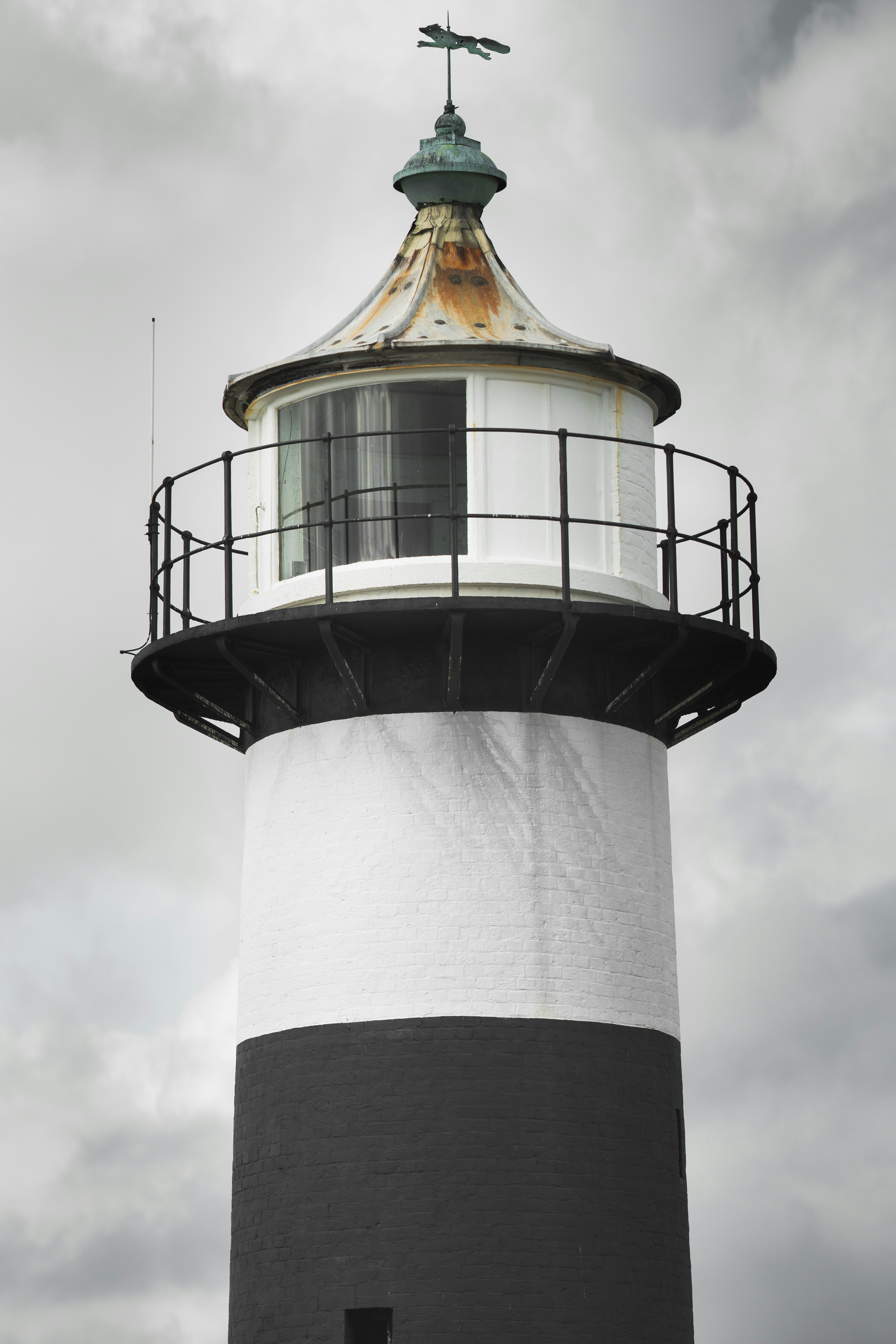 A classic lighthouse with a distinctive black and white pattern, featuring a copper roof and weather vane against a cloudy sky.