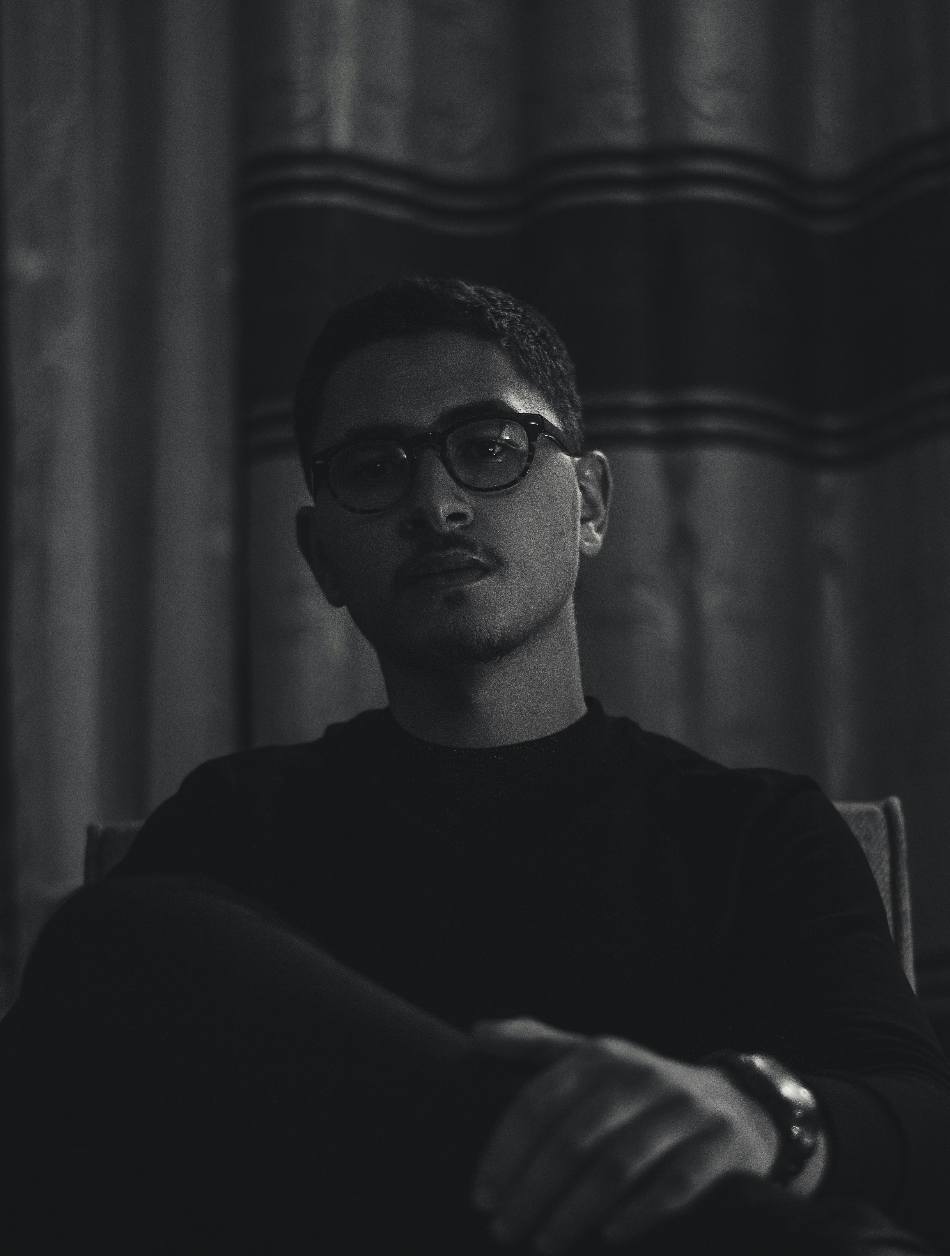 A thoughtful individual seated in a dimly lit room, framed by soft fabric curtains, exuding a sense of introspection.