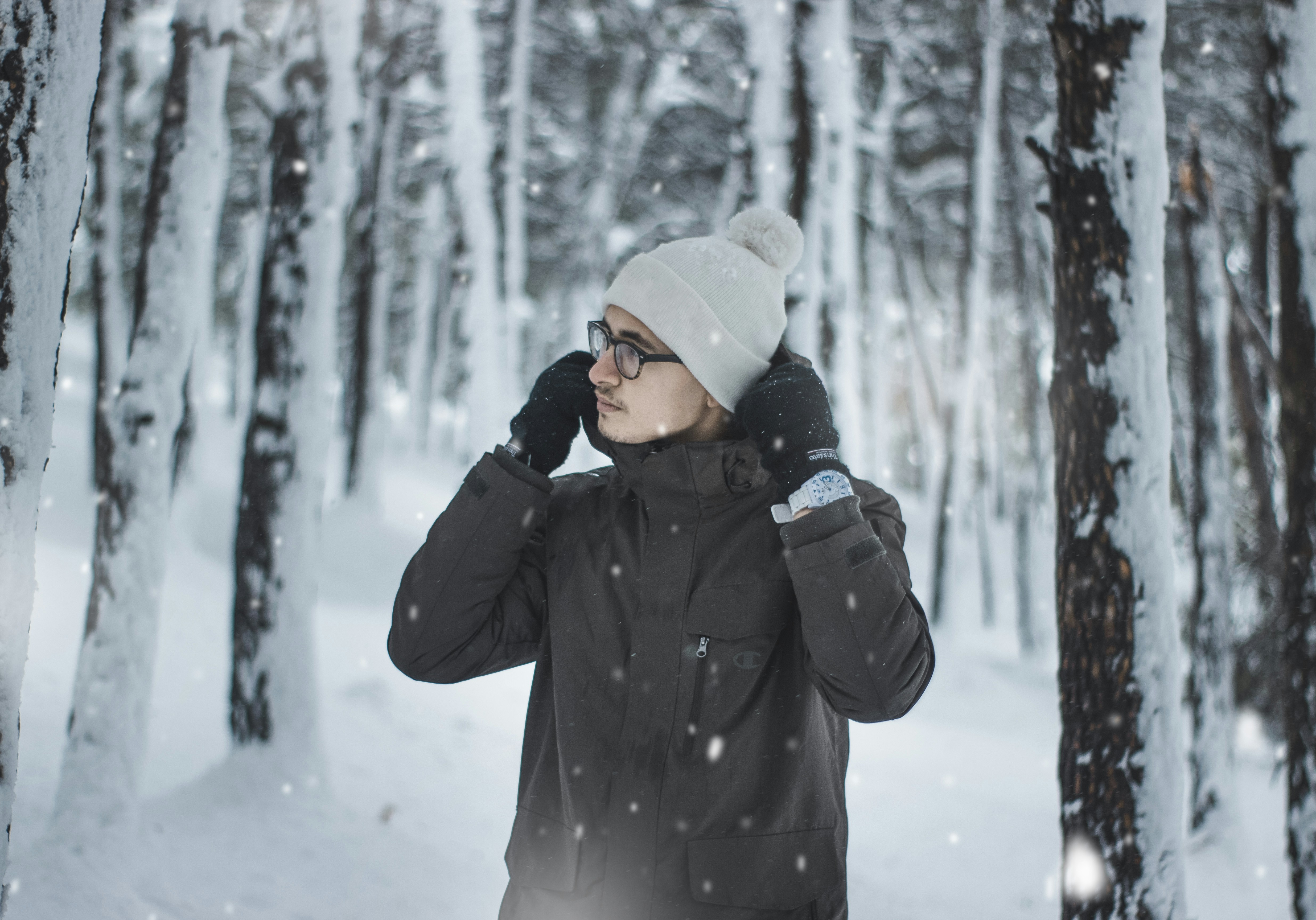 Person adjusting their hat amidst a serene snowy forest, surrounded by tall trees blanketed in white.