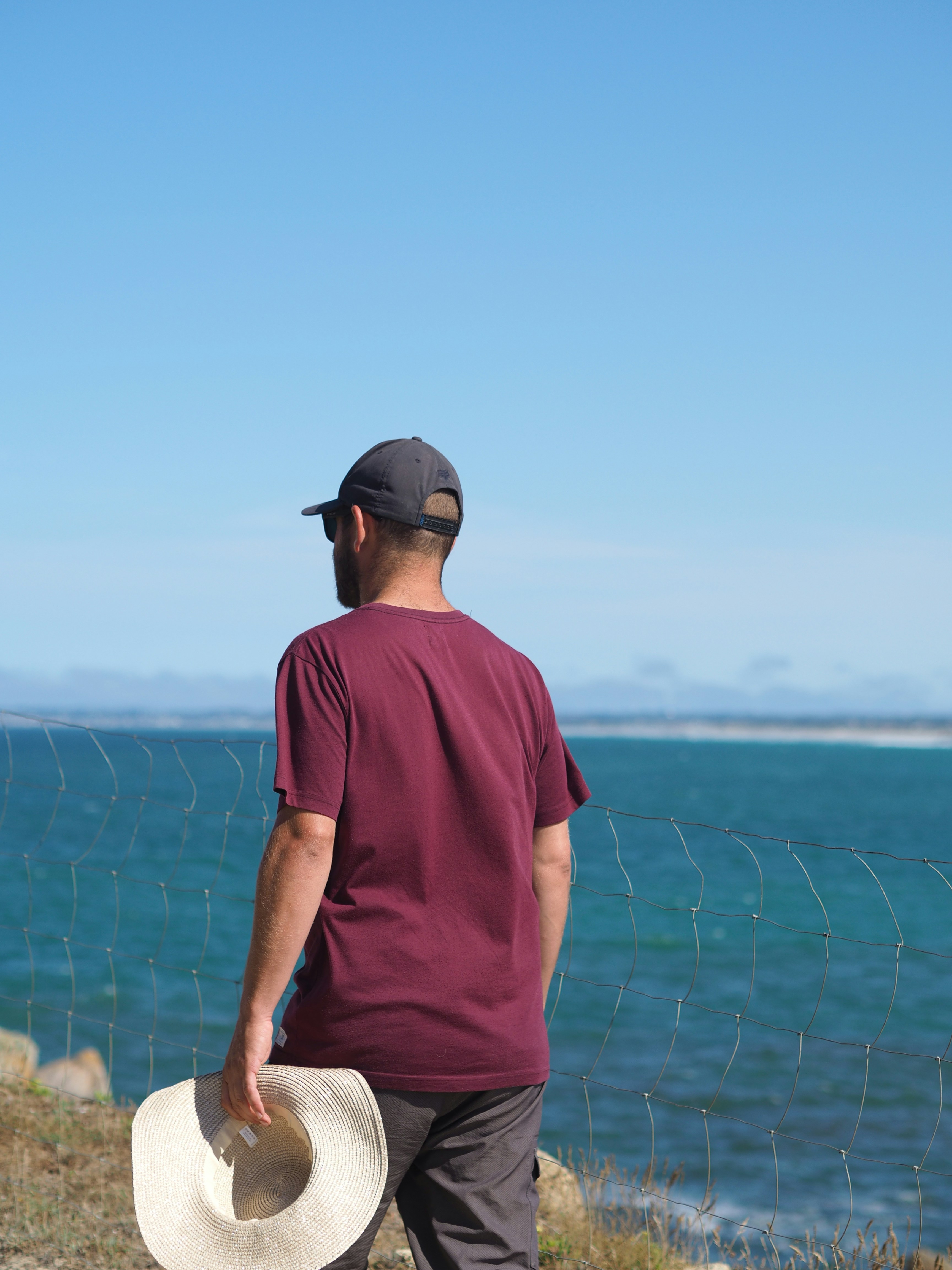 Man in a maroon shirt and hat stands by the ocean, holding a wide-brimmed hat, gazing at the horizon.