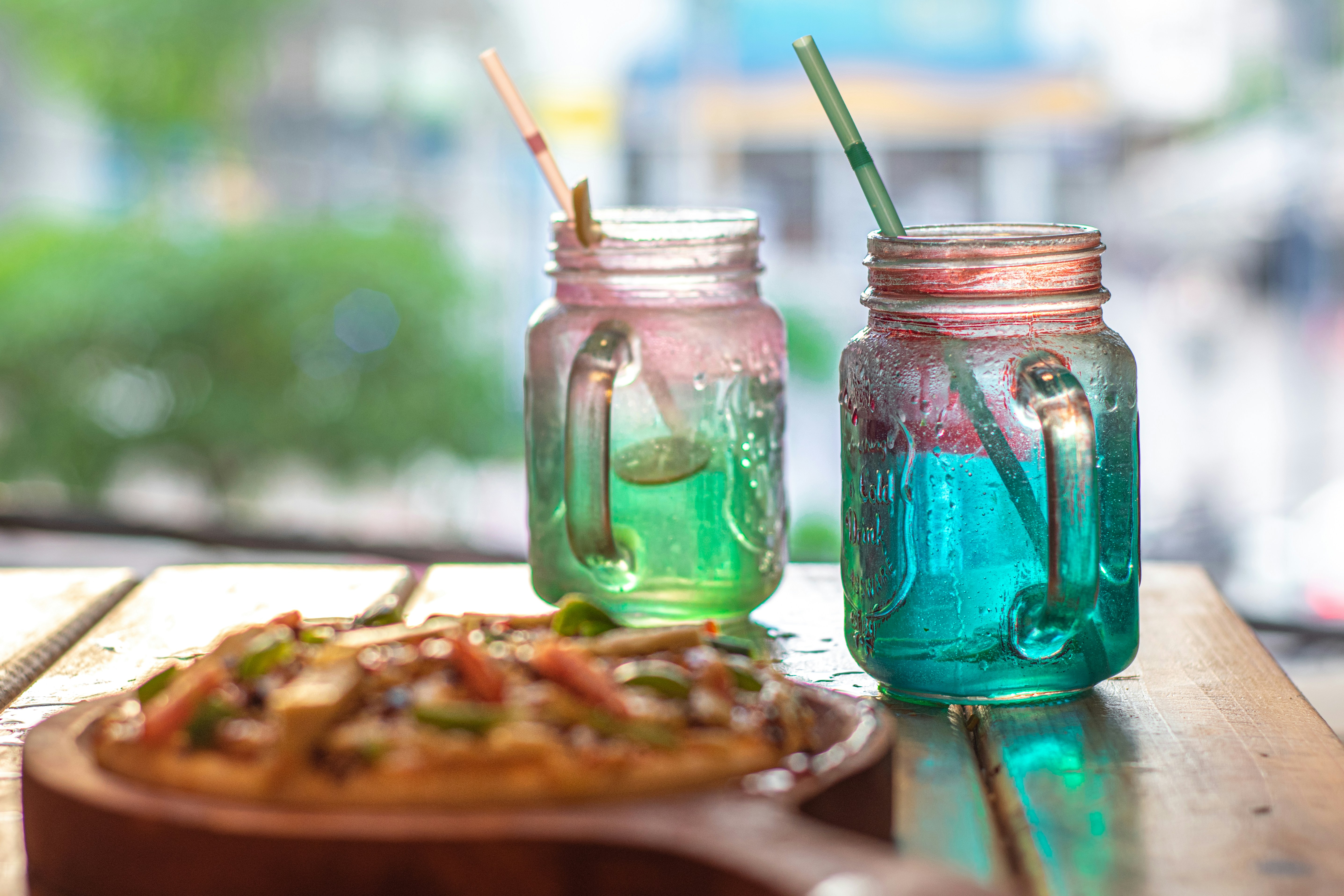 Two mason jars filled with vibrant blue and green drinks sit on a wooden table, with a blurred dish in the foreground. Straws in each jar add a playful touch.