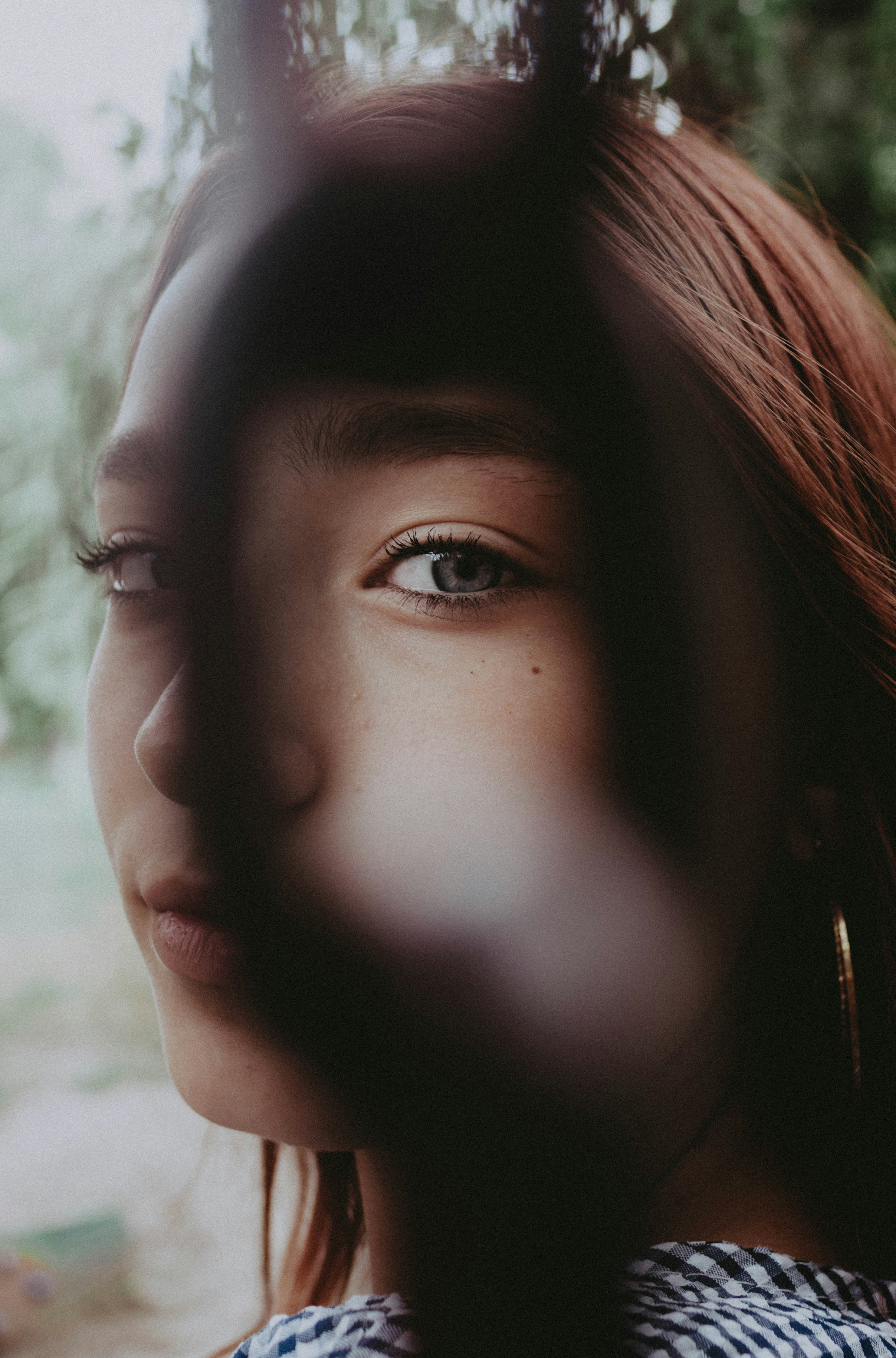 A young woman gazes thoughtfully through a natural frame, her expressive eyes revealing a story of contemplation. Soft focus enhances the intimate atmosphere.