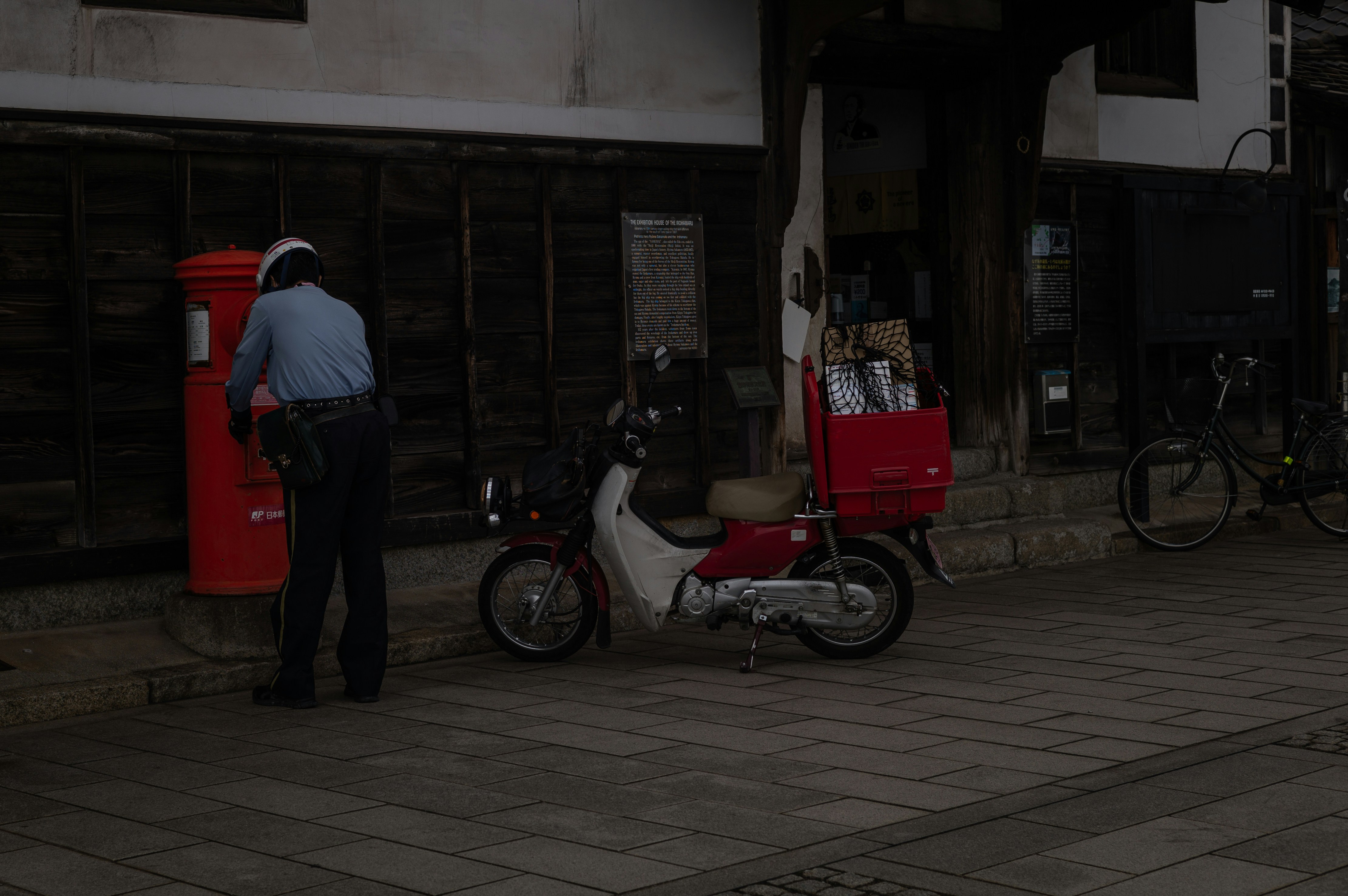 A postal worker interacts with a bright red mailbox beside a motorcycle in a quaint urban setting, showcasing daily life in a serene neighborhood.