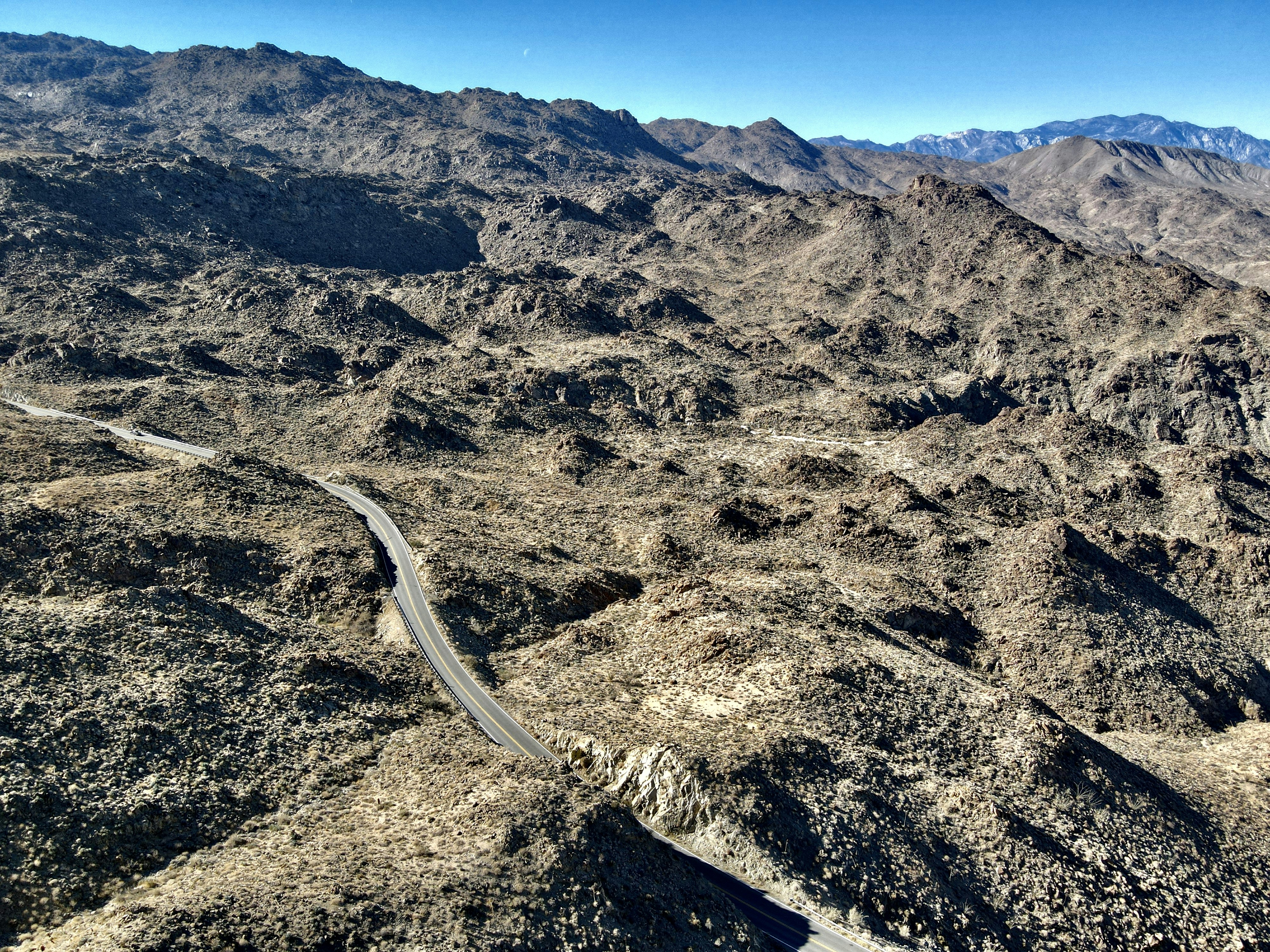 A winding road cutting through rugged, rocky desert mountains under a clear blue sky.