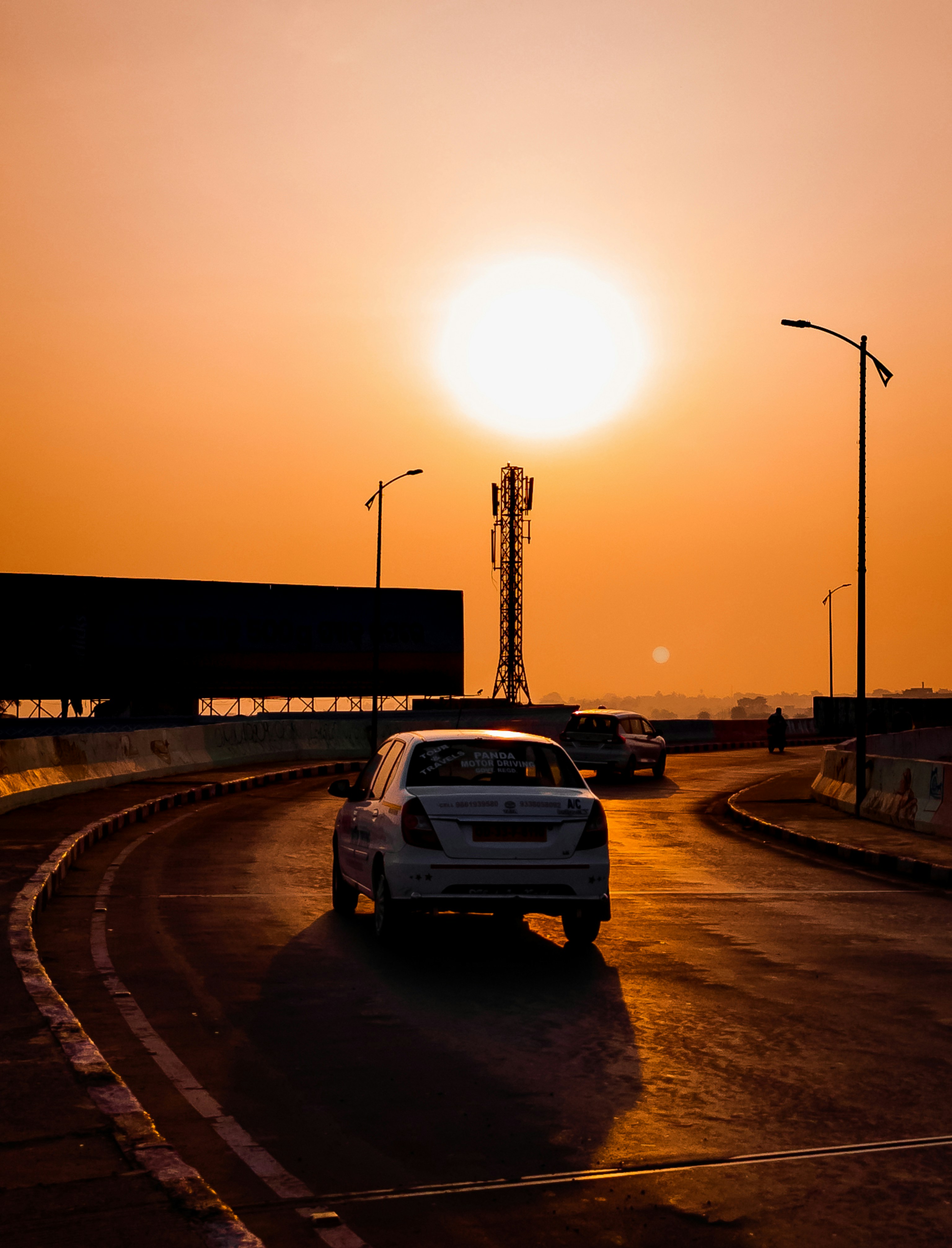 Curved highway bathed in warm orange sunset with passing cars. Urban silhouettes, streetlights, and a distant tower anchor the composition.