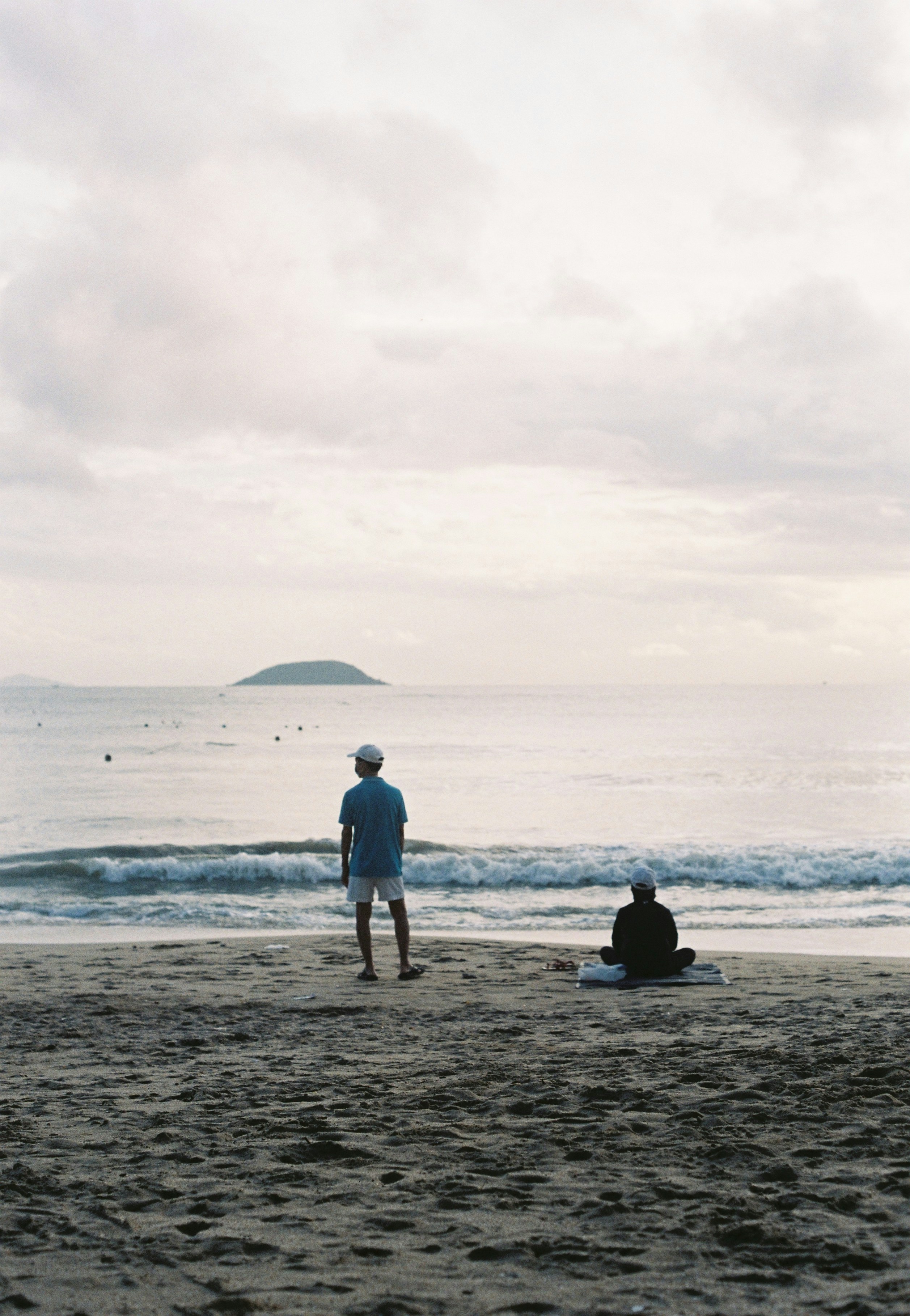 Two figures on a sandy beach, one standing and gazing at the ocean while the other sits quietly, with a distant island visible on the horizon.