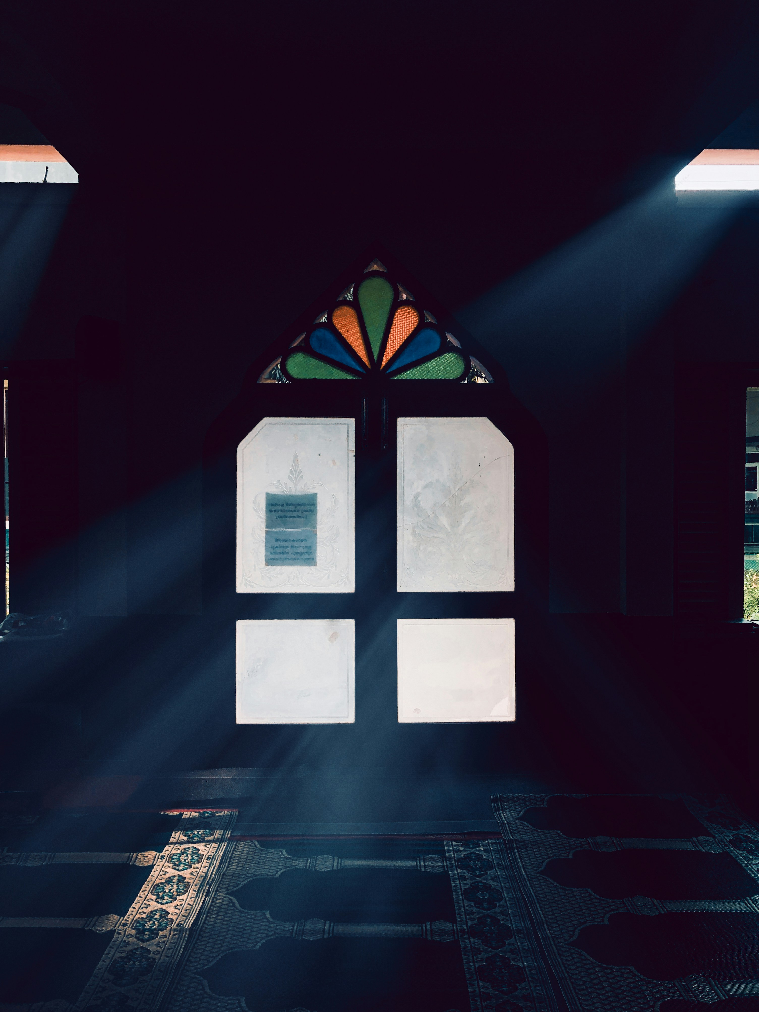 Light beams filtering through a stained glass window, casting an ethereal glow in a dimly lit space. The intricate design of the glass adds a touch of color to the serene atmosphere.