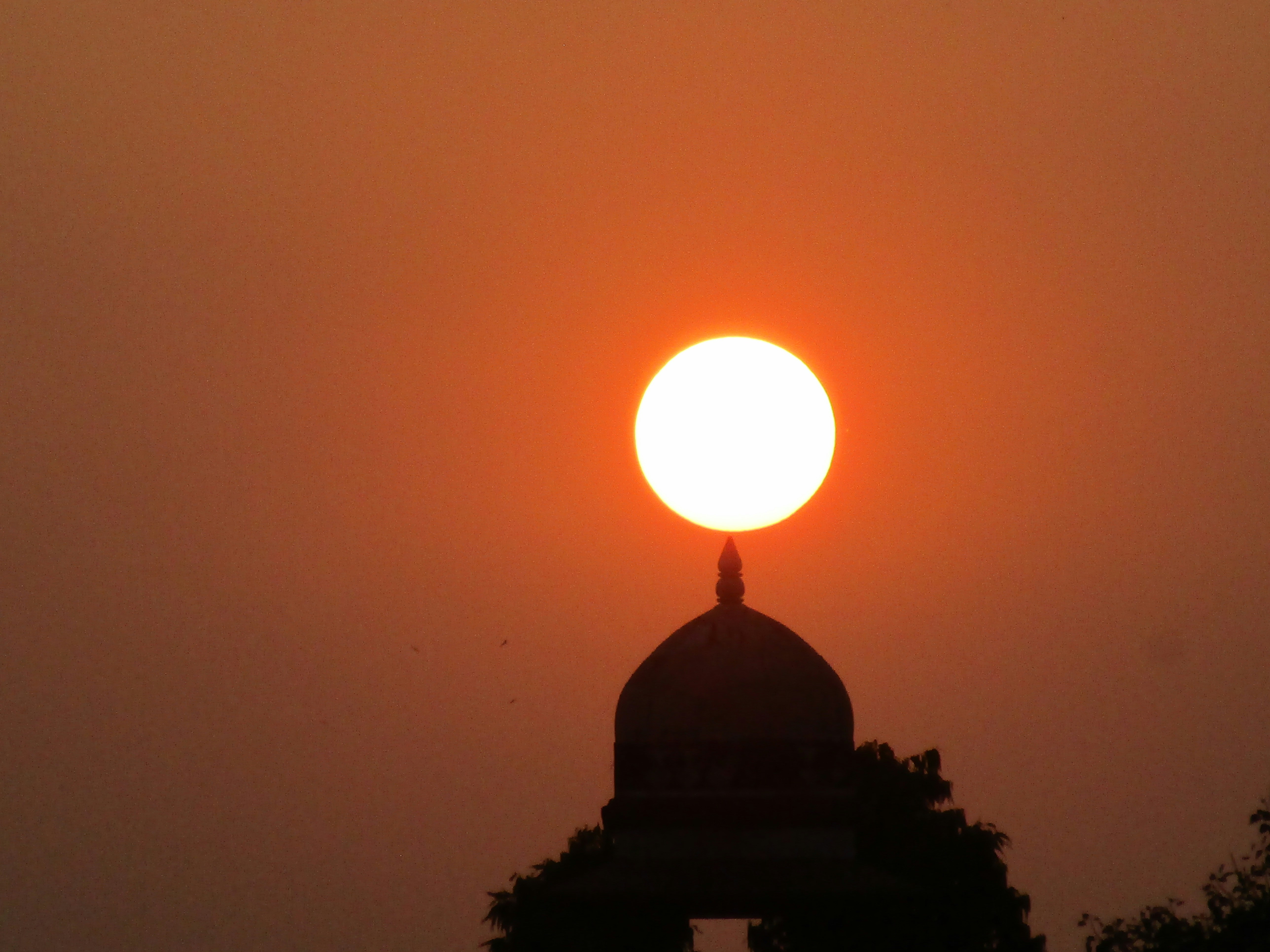 Soft amber sunset with a silhouetted domed structure and a bright sun centered above its peak; warm gradient sky and subtle foreground silhouettes.