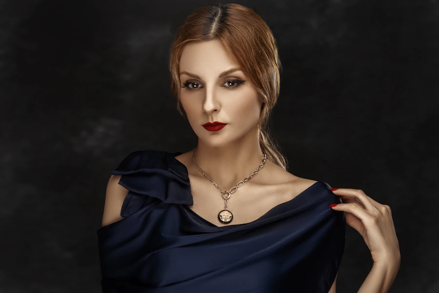 Elegant studio portrait of a woman with red lipstick, pulled-back hair, and a dark blue draped dress against a black background. She wears a pendant necklace and rests one hand on the fabric.