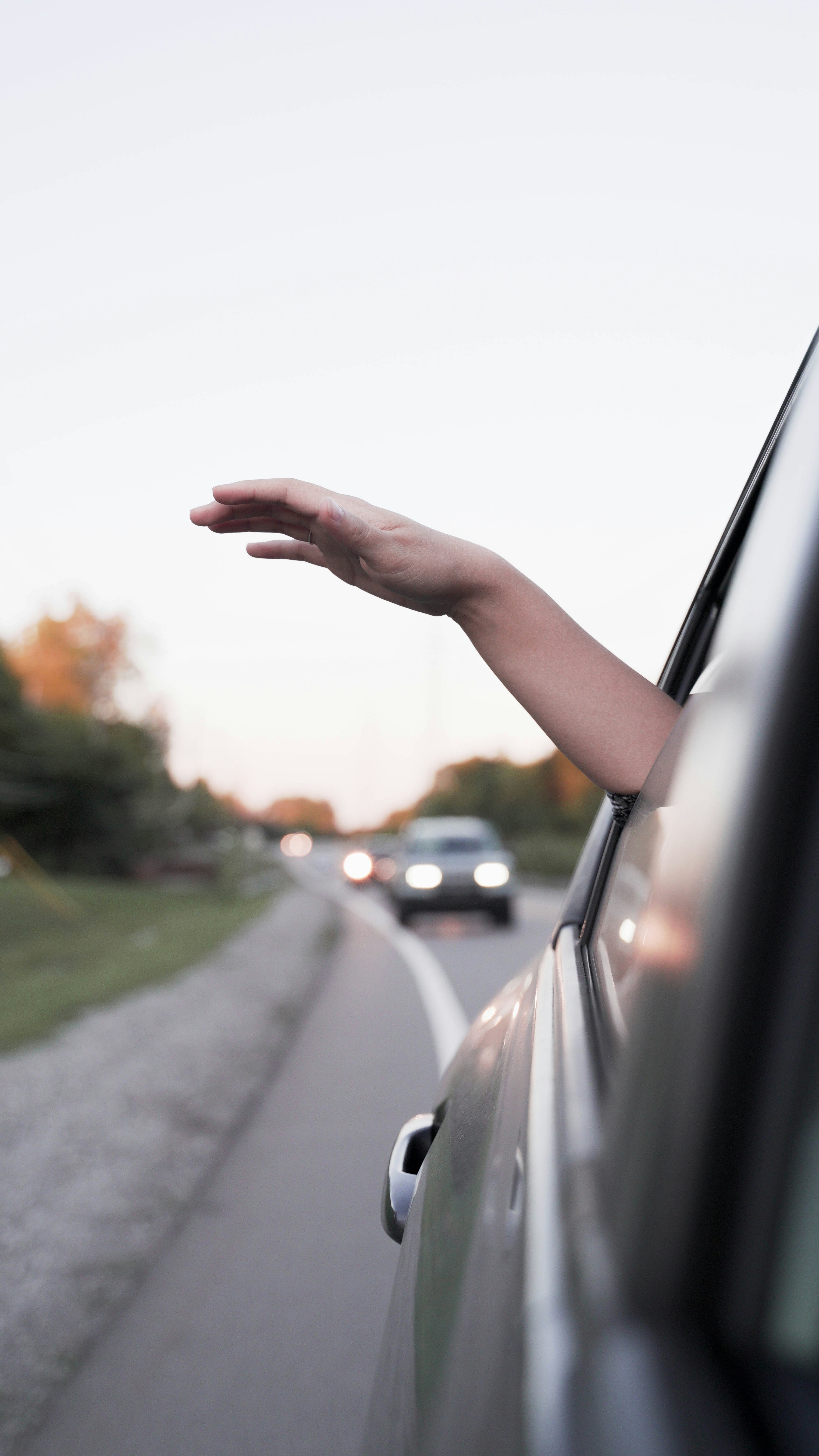 Hand reaching out of a car window against a backdrop of a winding road and soft sunset hues.