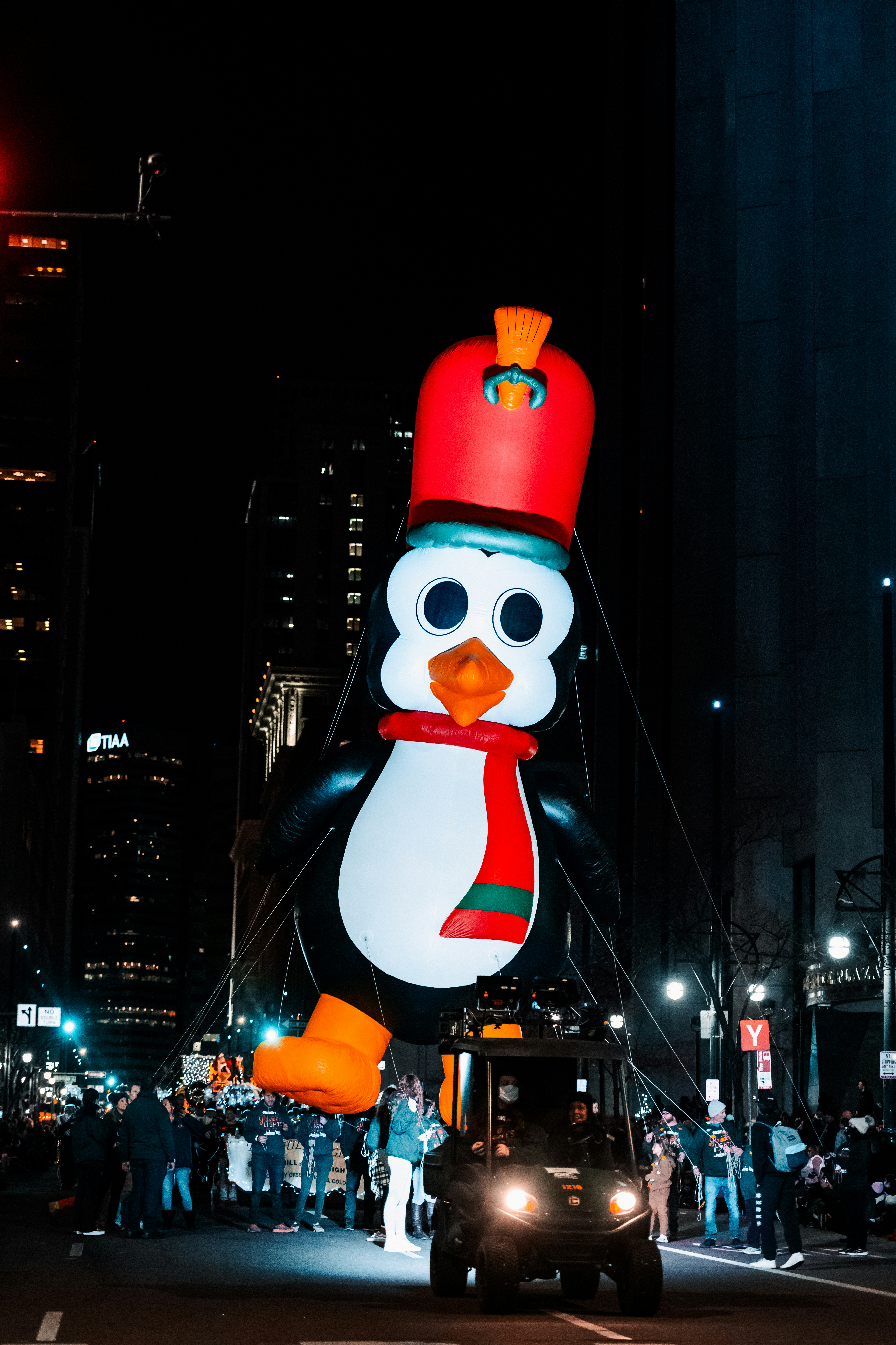 Giant inflatable penguin with a festive hat floats above a crowd during a nighttime parade. Bright city lights create a lively atmosphere.