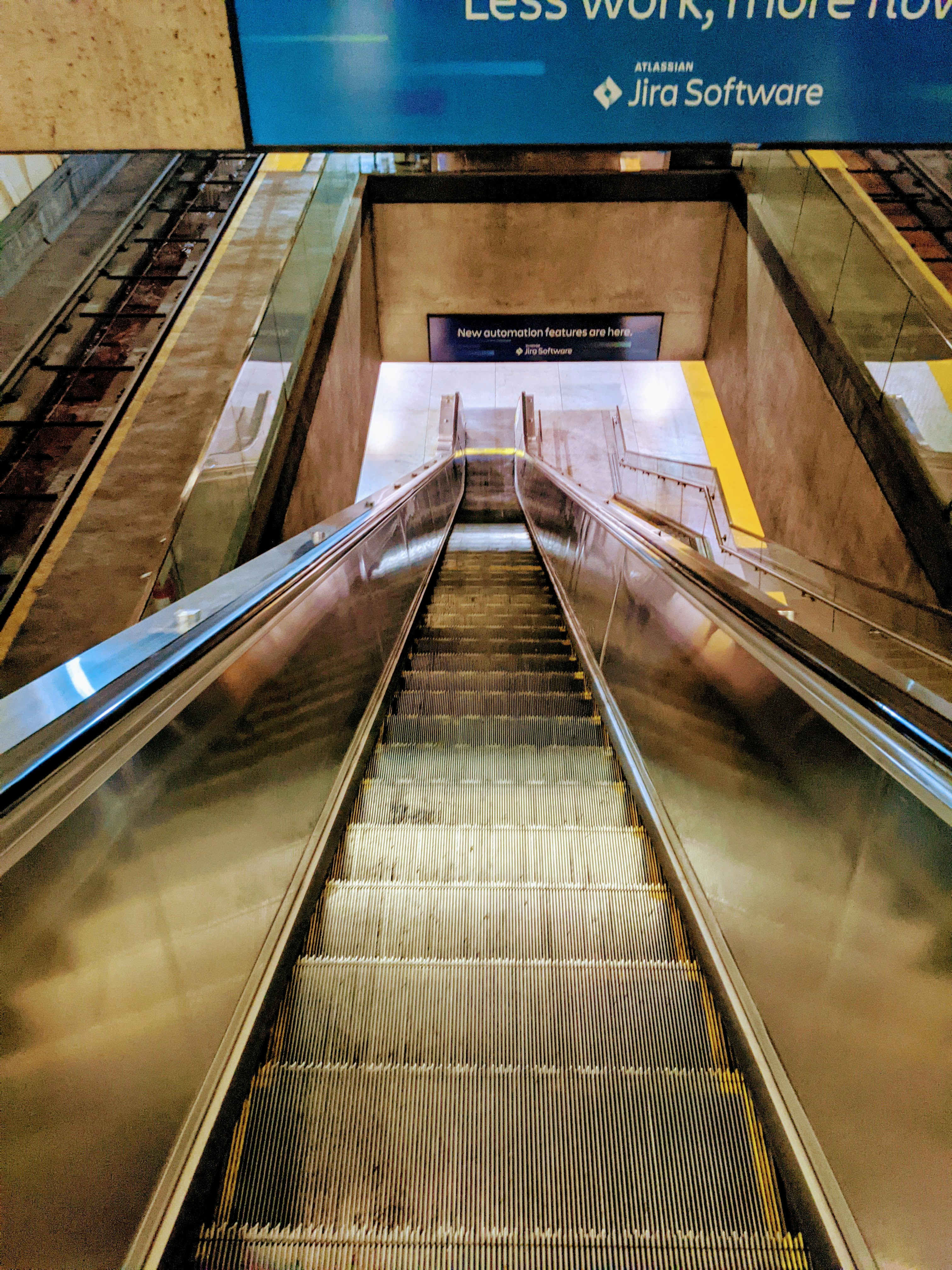 Escalator leading down into a modern transit area, framed by concrete walls and illuminated signage above. The design emphasizes functionality and urban efficiency.