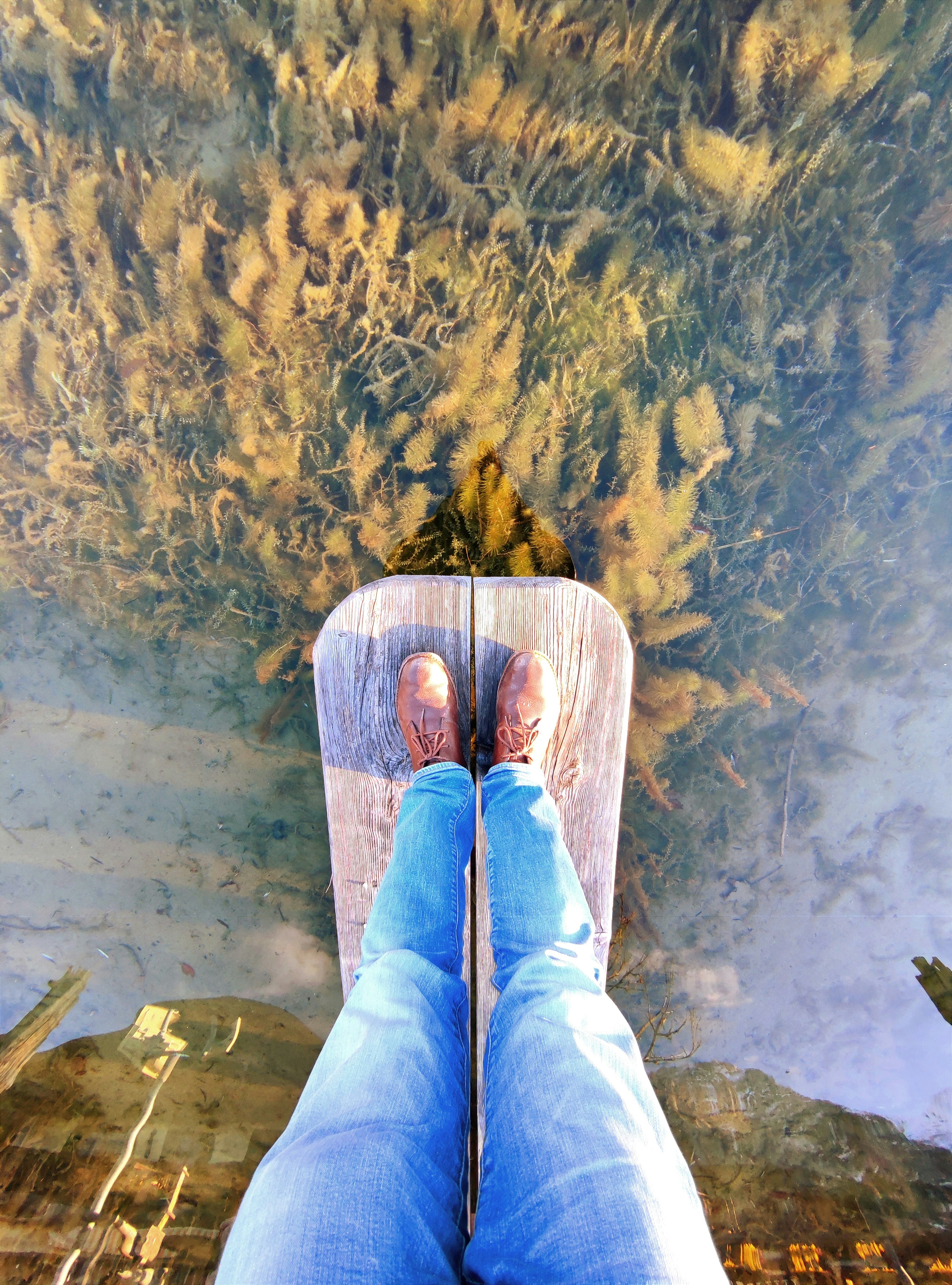 Top-down photograph of a person standing on a weathered wooden plank extending over calm, glassy water; blue jeans and brown shoes anchor the frame. The composition emphasizes surface reflections and quiet surroundings.