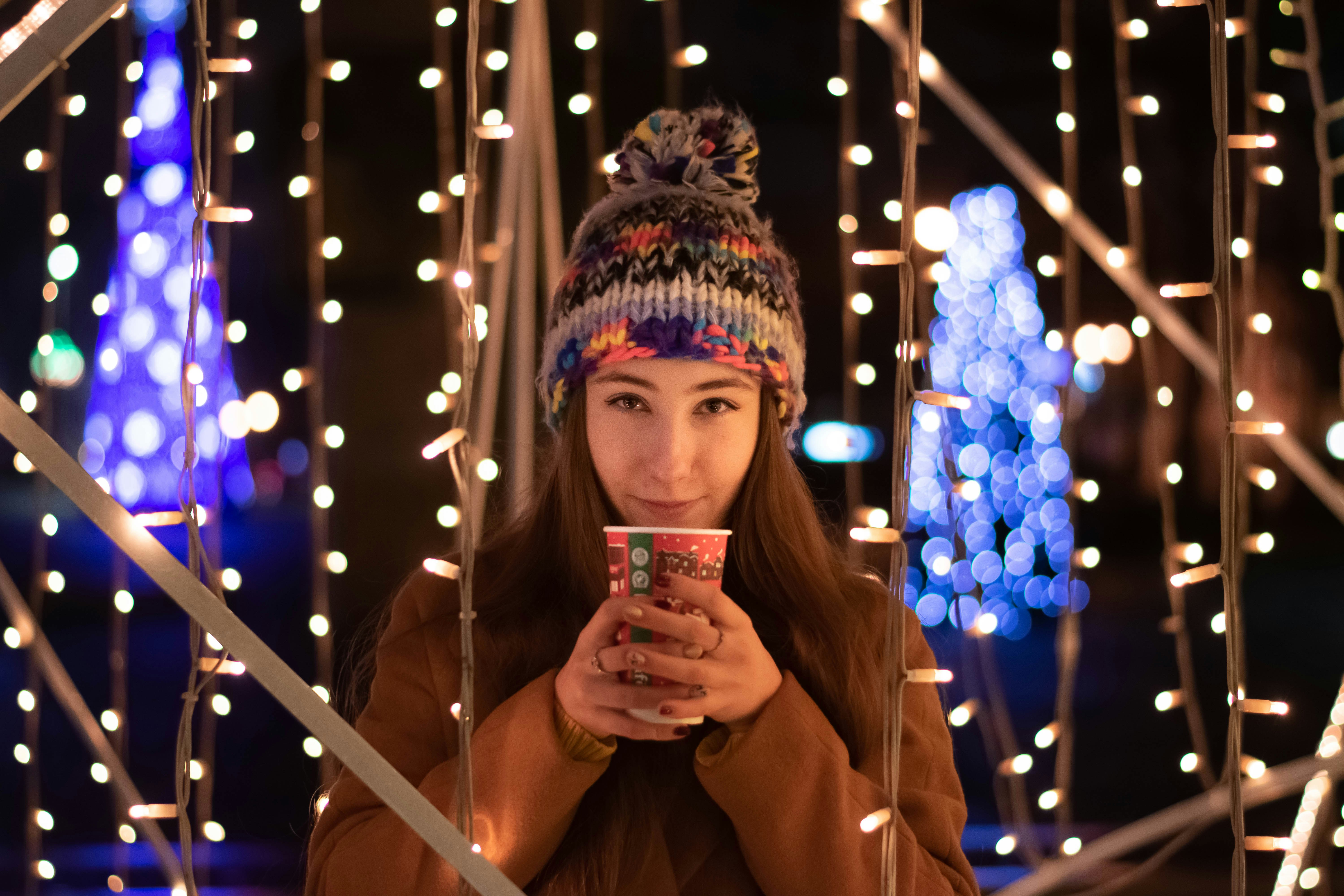 Person holding a cup of hot beverage, surrounded by festive string lights and wearing a colorful knit hat.