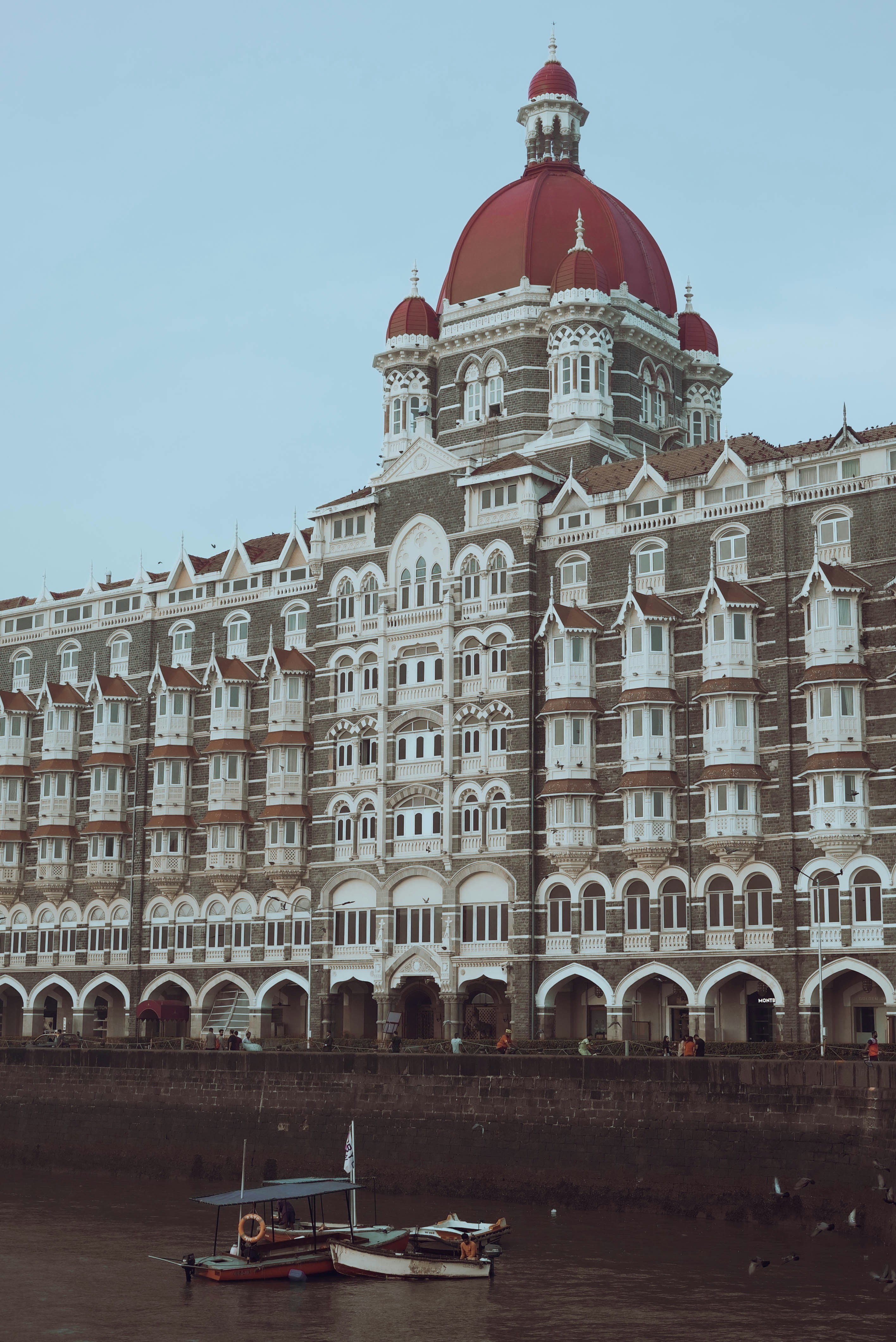 The iconic Taj Mahal Palace hotel stands majestically by the waterfront, showcasing its distinct red dome and intricate architectural details. A small boat rests in the foreground, adding to the scene's charm.