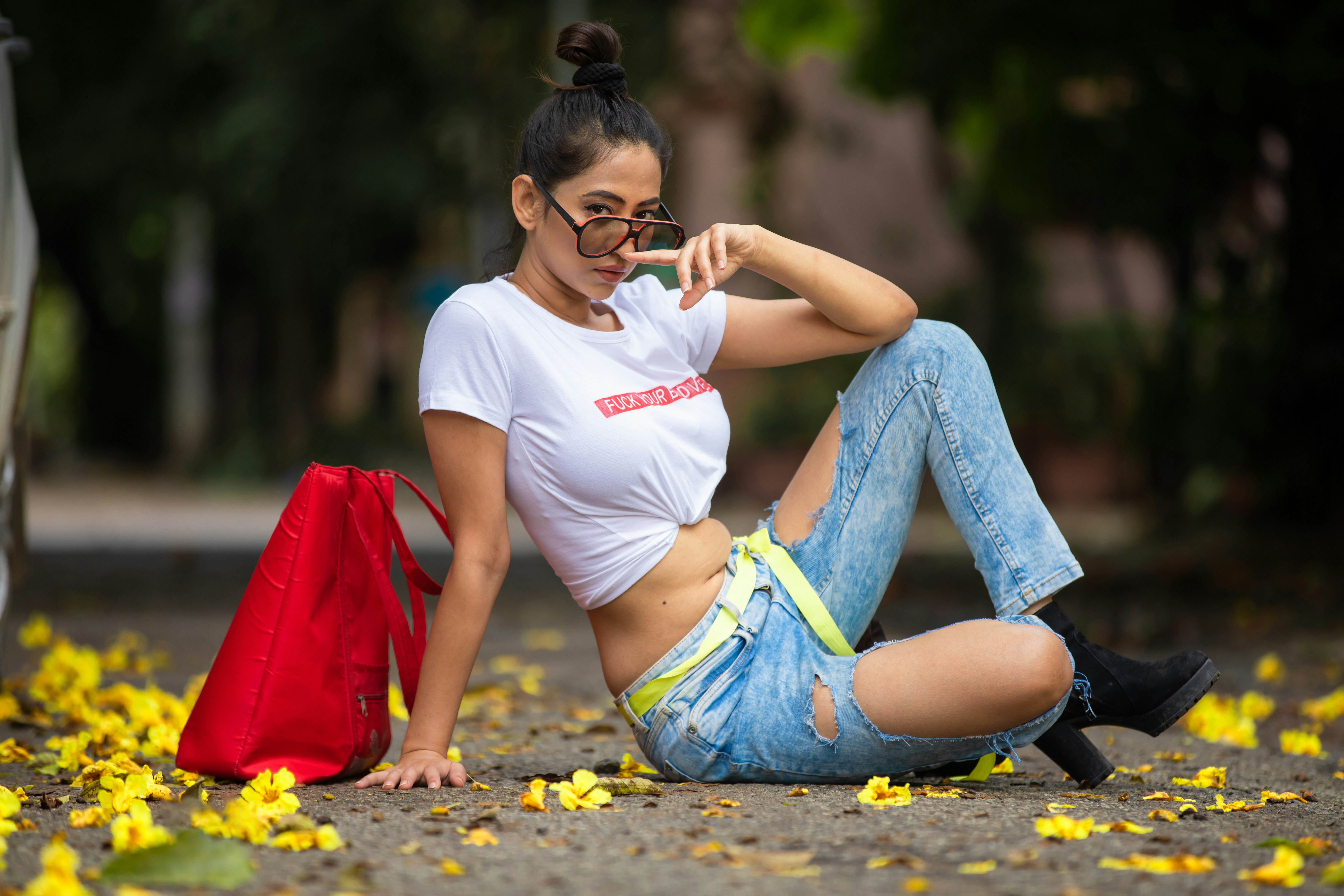 Woman in casual attire sitting on a street surrounded by yellow flowers, with a vibrant red bag.