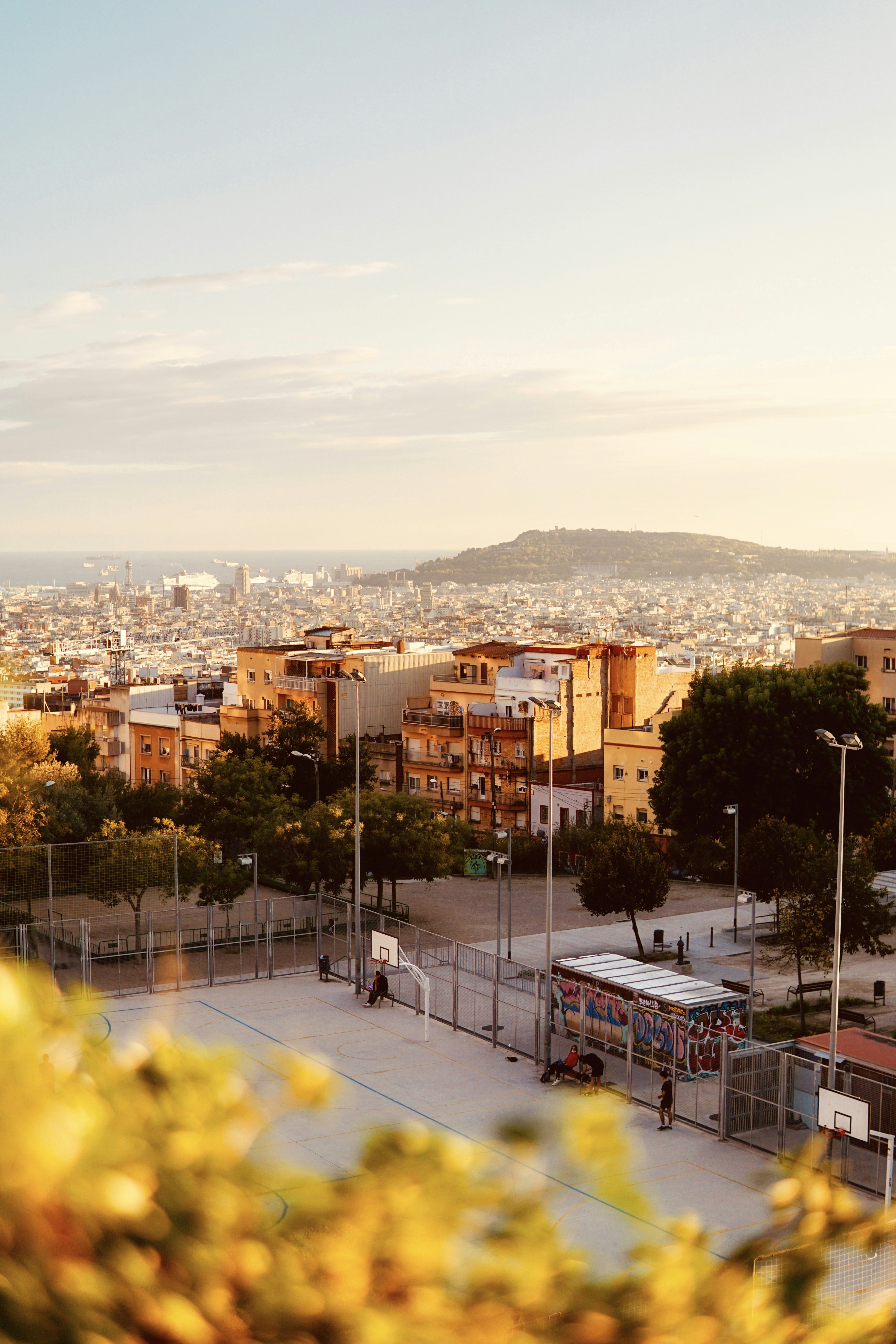 View of a vibrant basketball court in Barcelona, framed by colorful buildings and distant hills under a soft sunset glow.