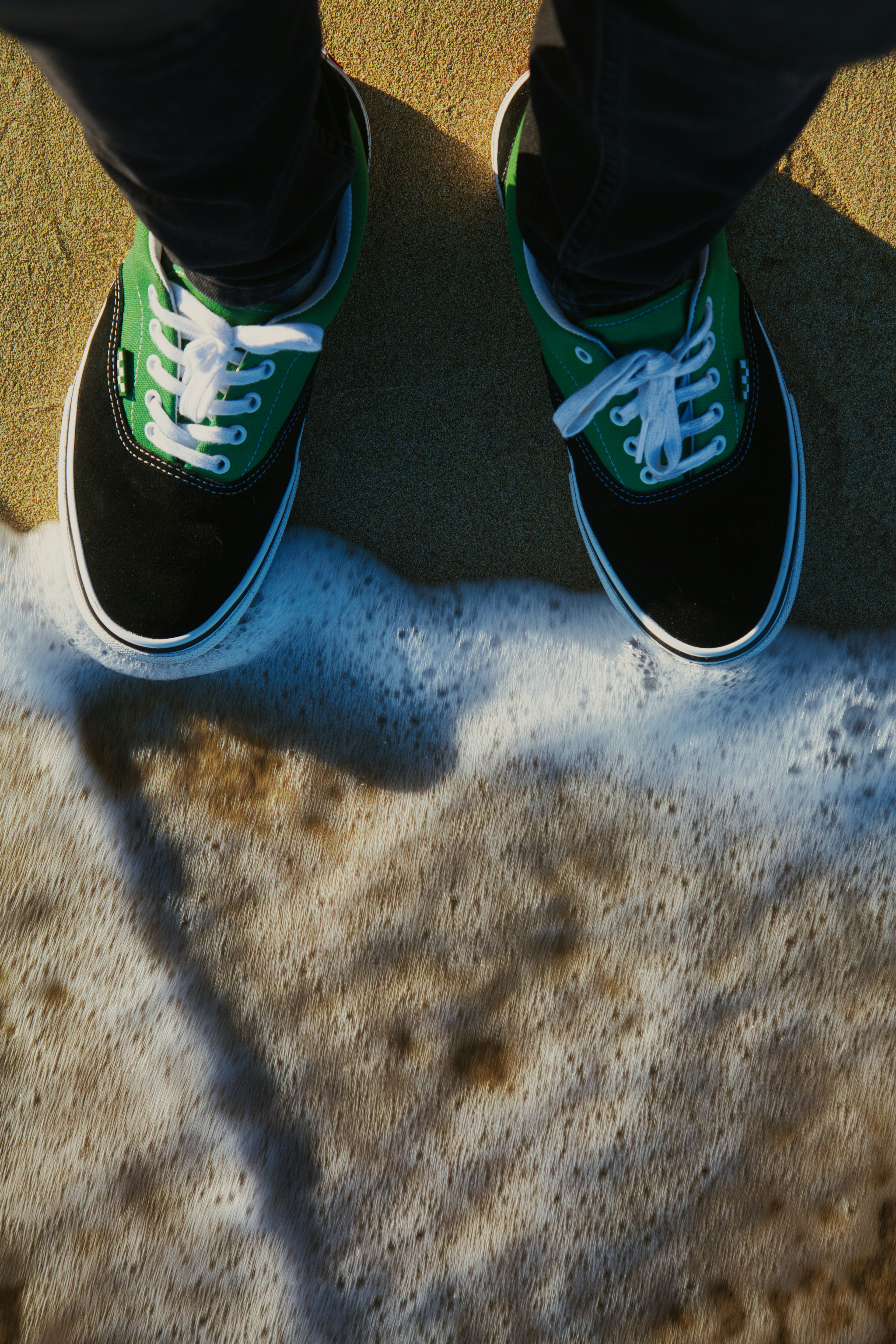 Feet in stylish sneakers resting on wet sand as ocean waves gently lap at the shore.