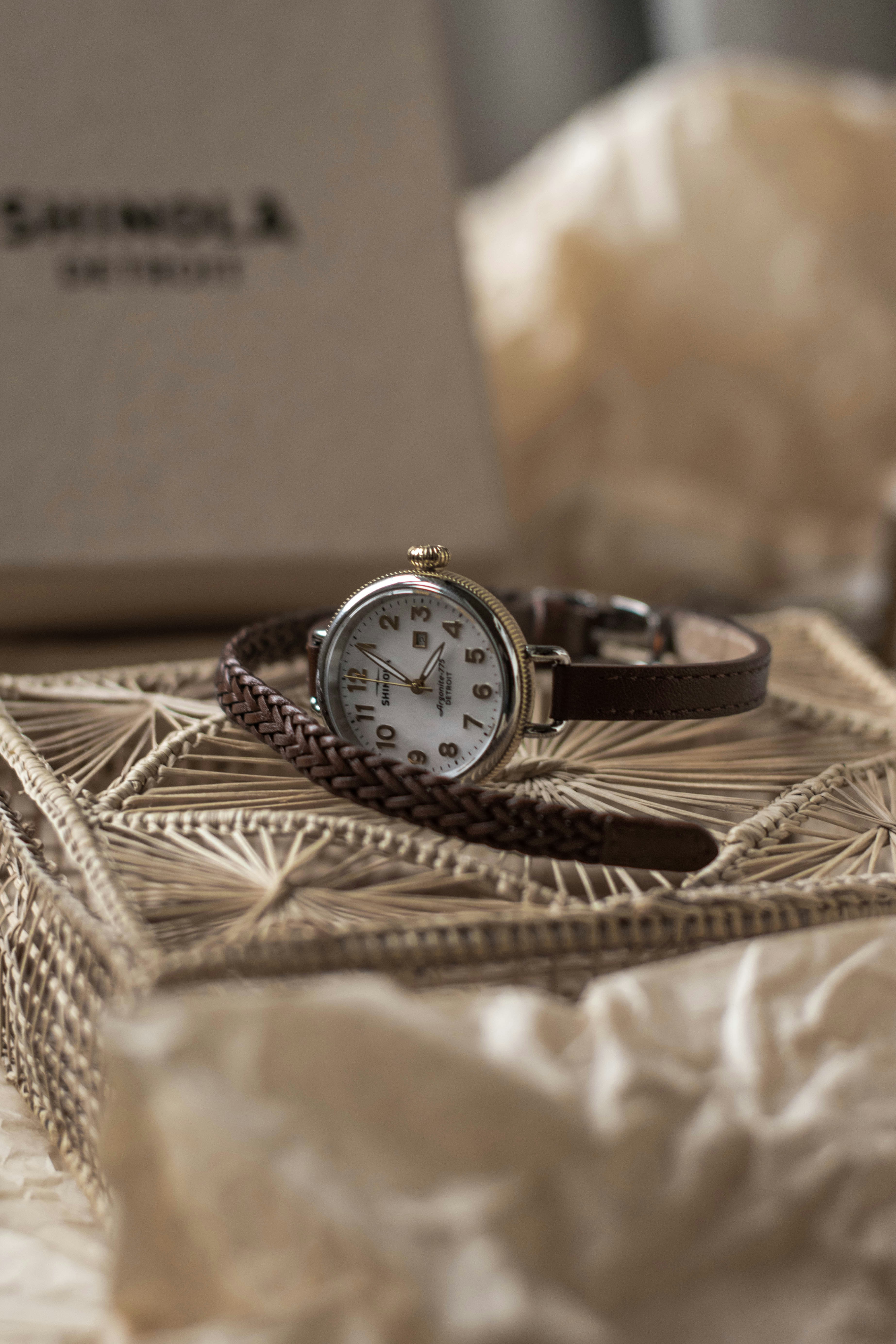 Classic wristwatch with a braided leather strap resting on a textured surface, accompanied by a subtle background of crumpled paper and a branded box.