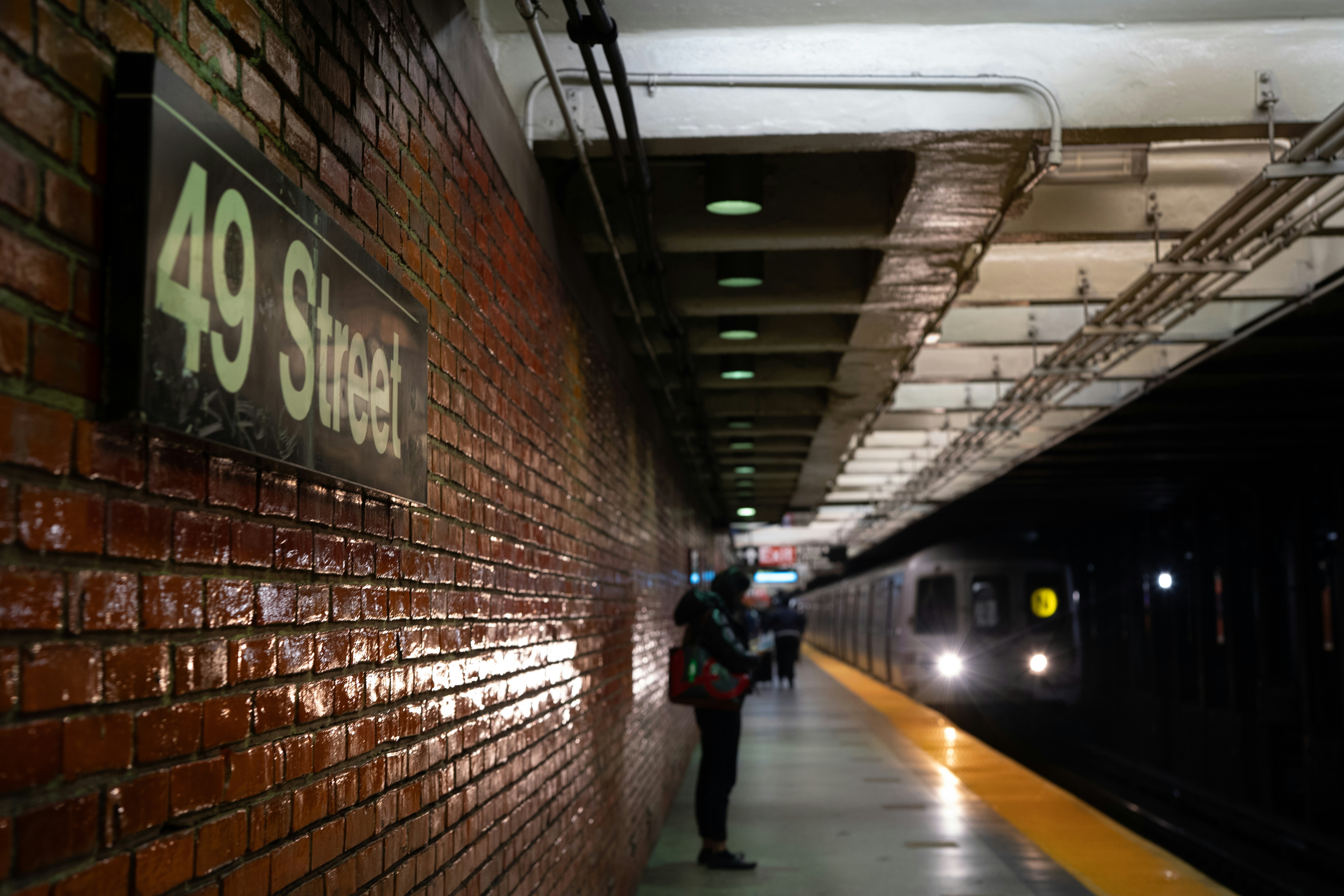 Subway station platform featuring a wet brick wall and a lone figure waiting for the train. The illuminated sign reads '49 Street.'