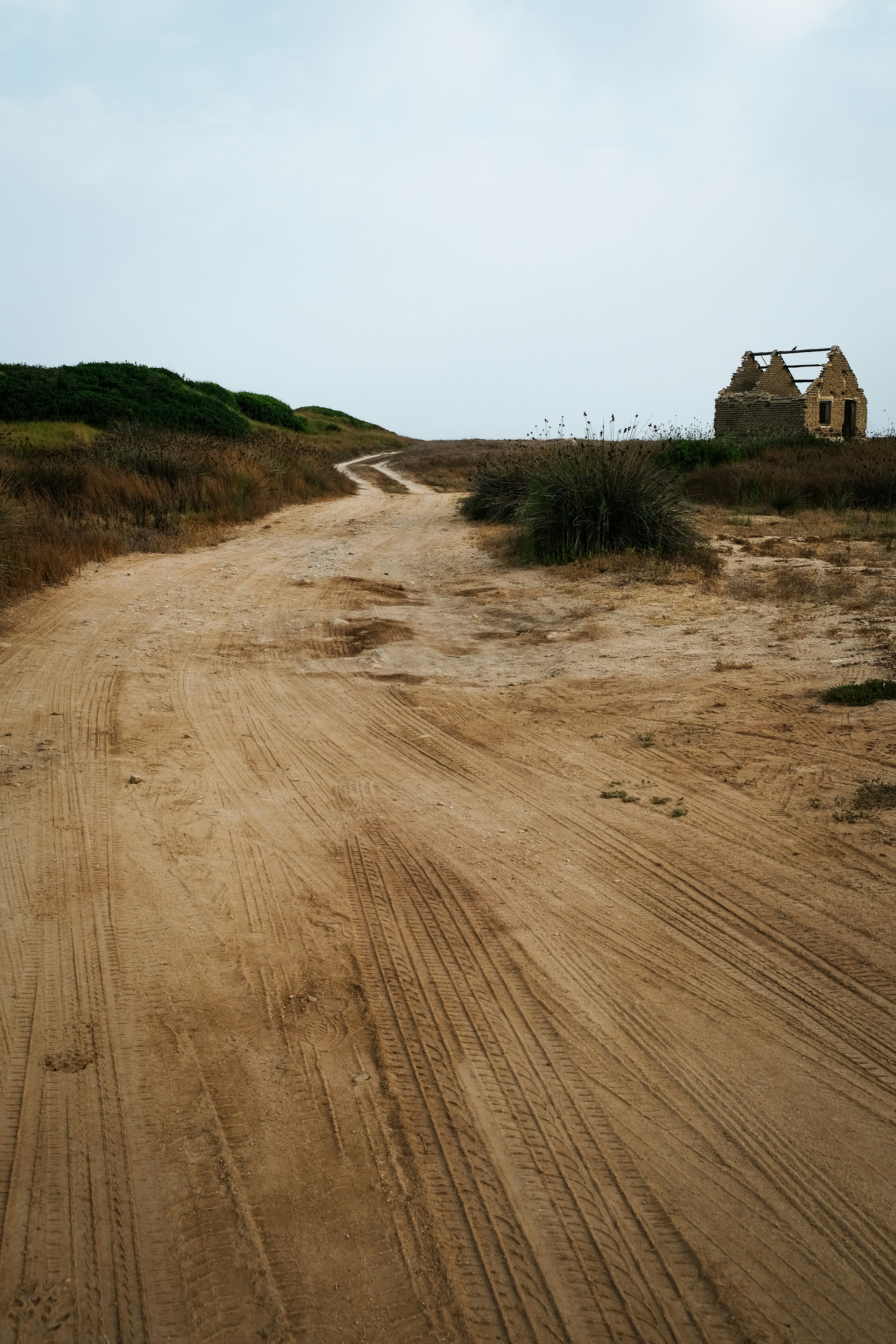 Winding dirt road leads to a dilapidated structure surrounded by overgrown grass under a cloudy sky.