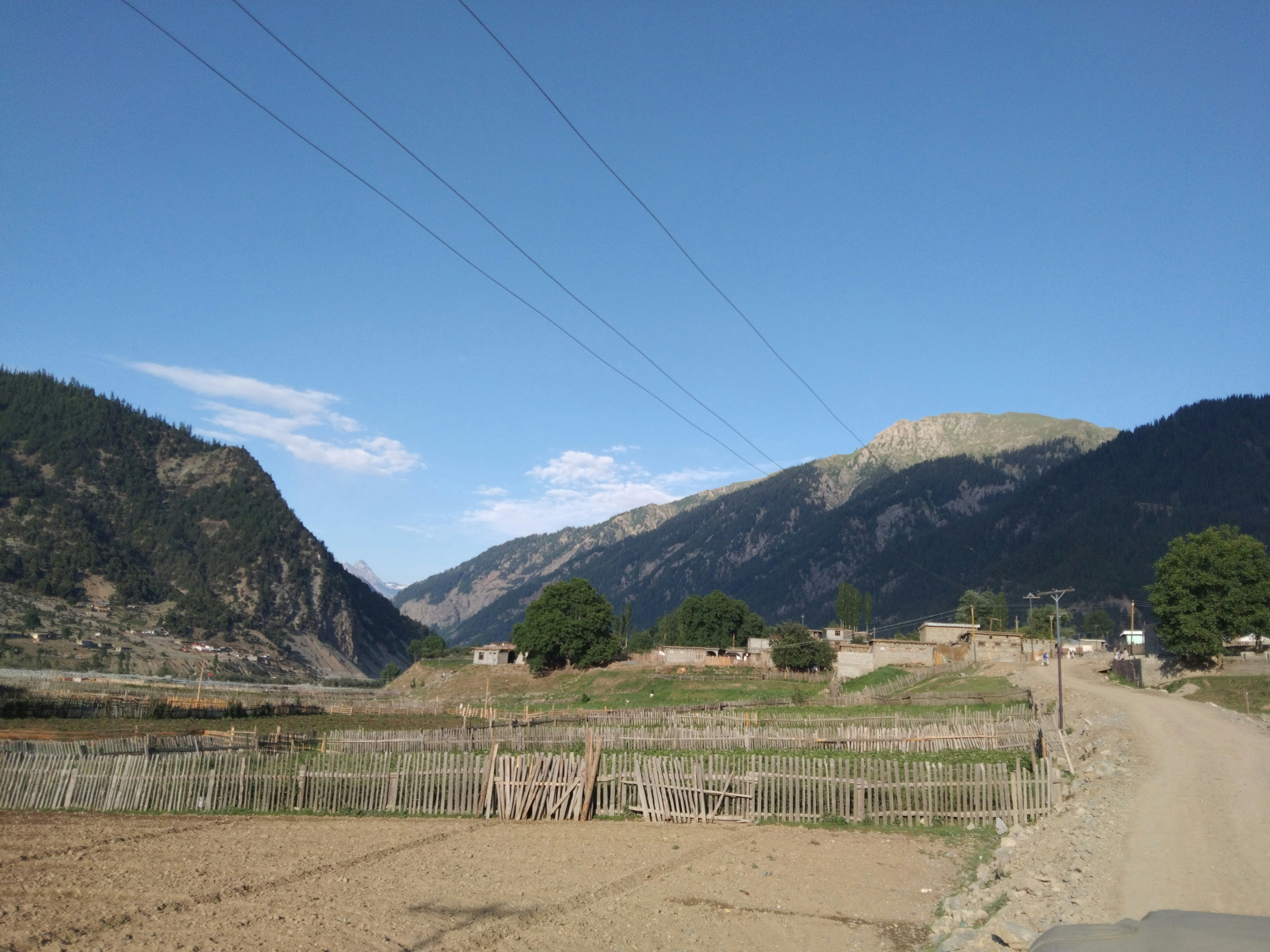 Landscape photograph of a rural field with a dirt road, wooden fence, and distant mountains under a clear blue sky.
