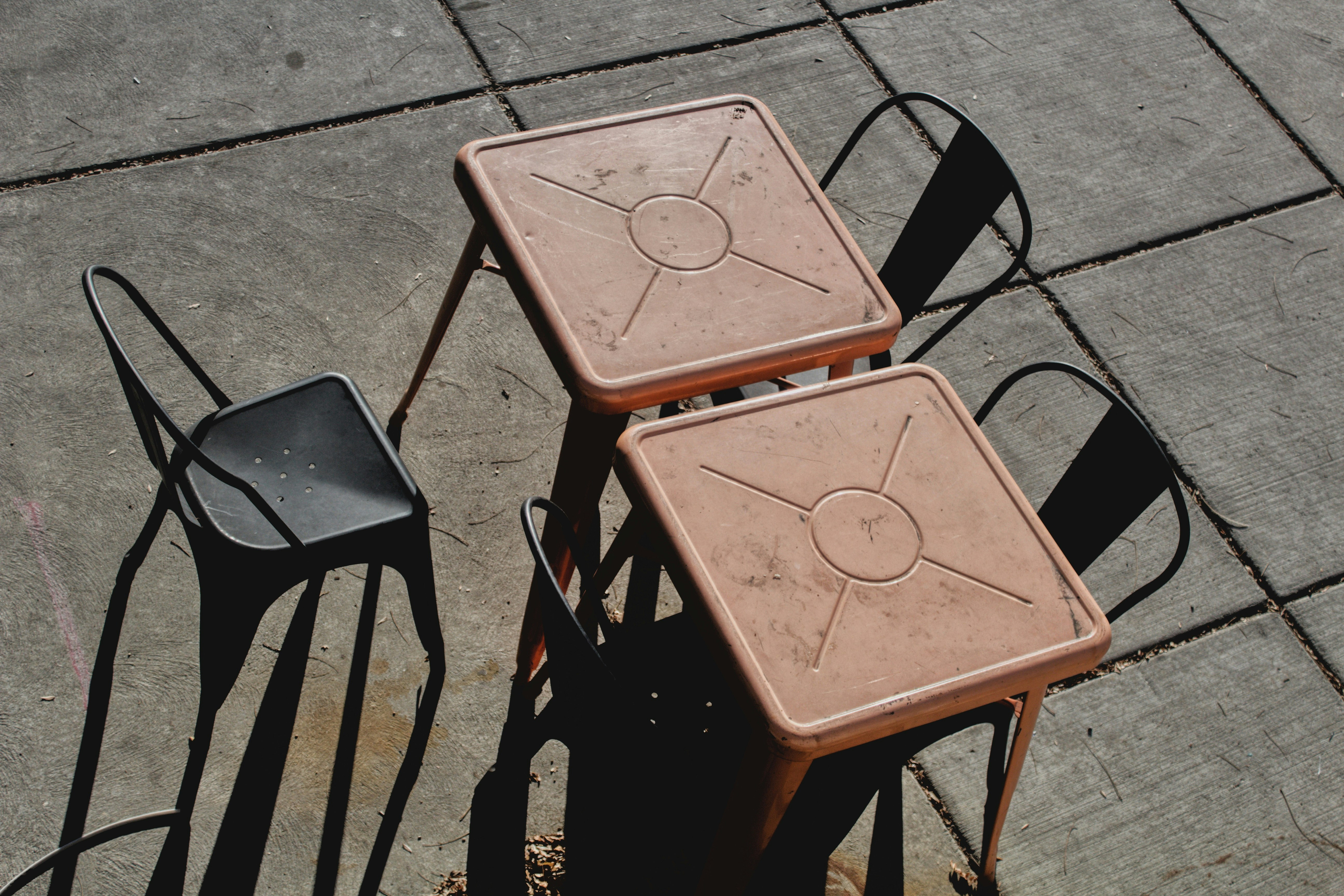Two tables with contrasting chairs cast intriguing shadows on a concrete surface, showcasing an interplay of geometry and urban life.