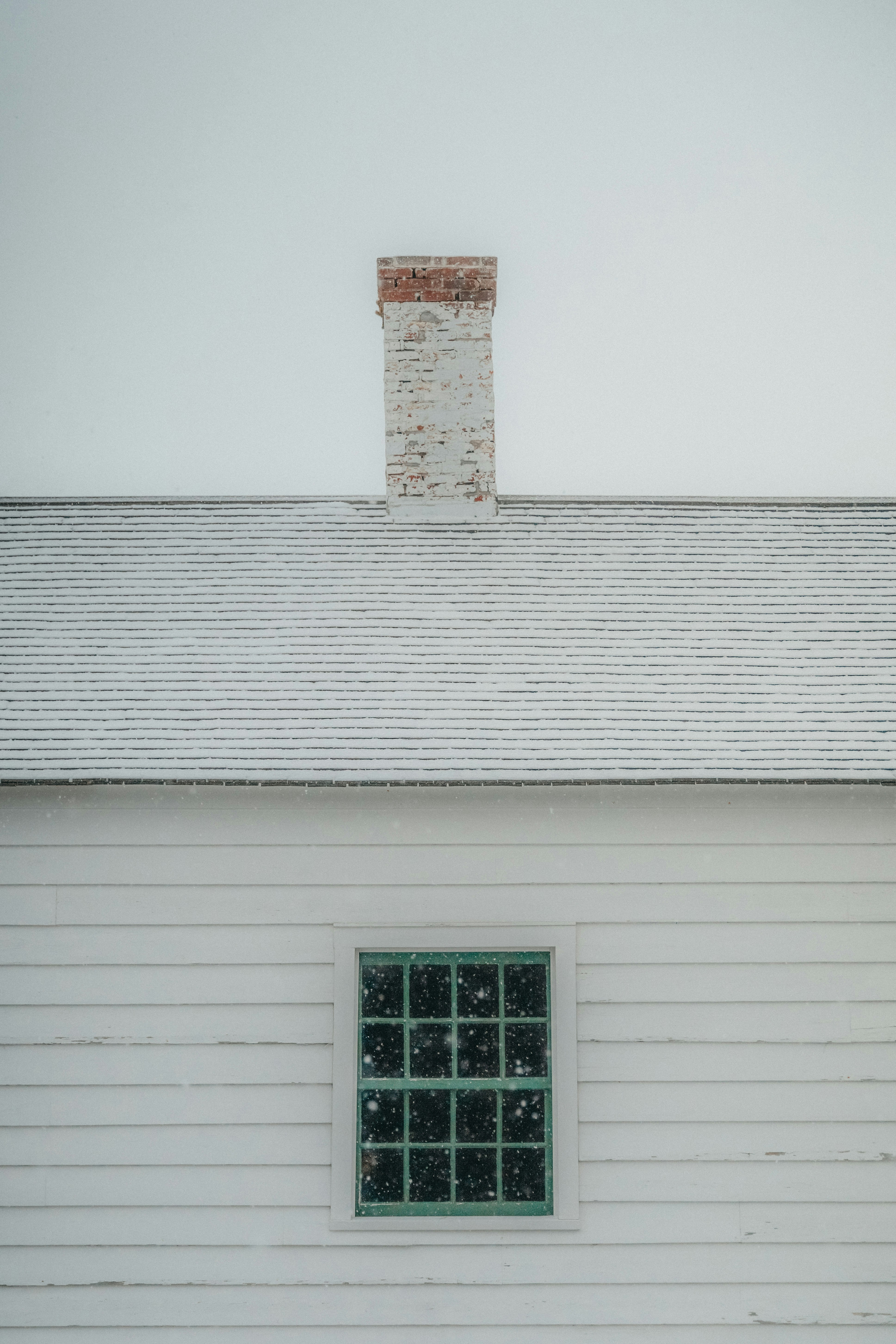 Snow-covered roof with a brick chimney and a green-framed window, set against a pale sky. The scene evokes a serene winter atmosphere.