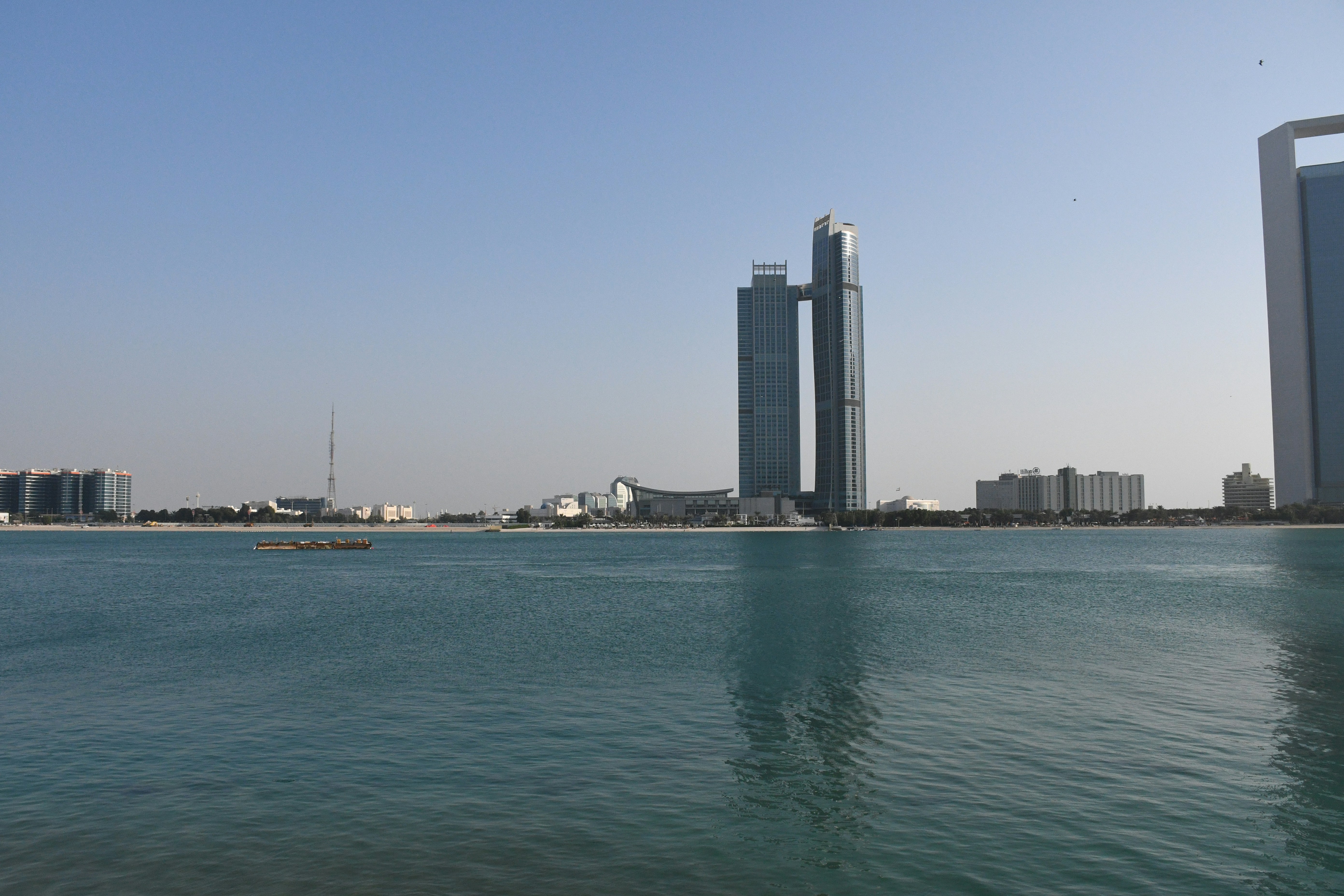 Modern skyscrapers reflecting in tranquil waters under a clear sky.