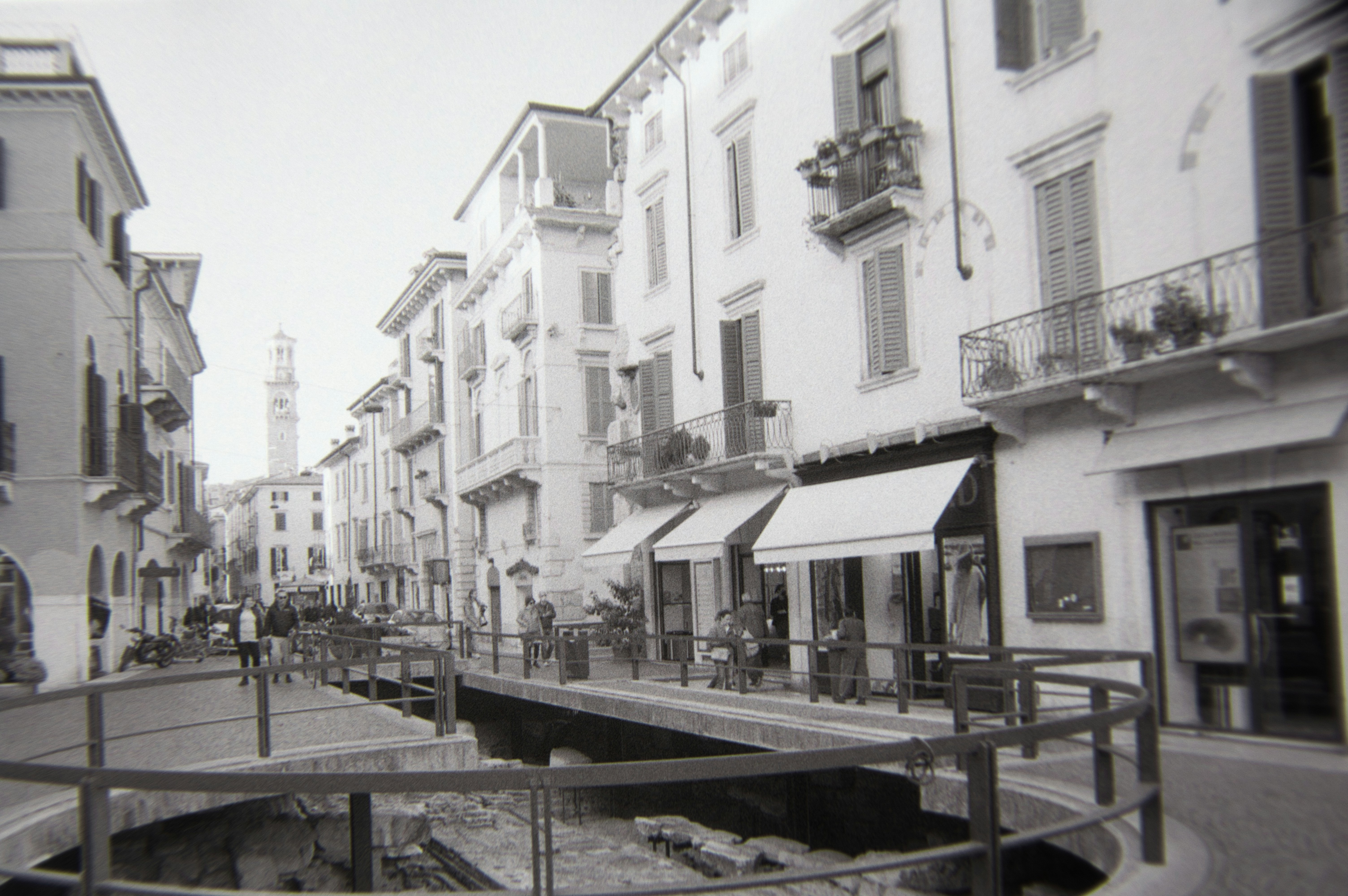 Black and white view of a Verona street with Roman excavations beneath, featuring classic architecture and pedestrians.