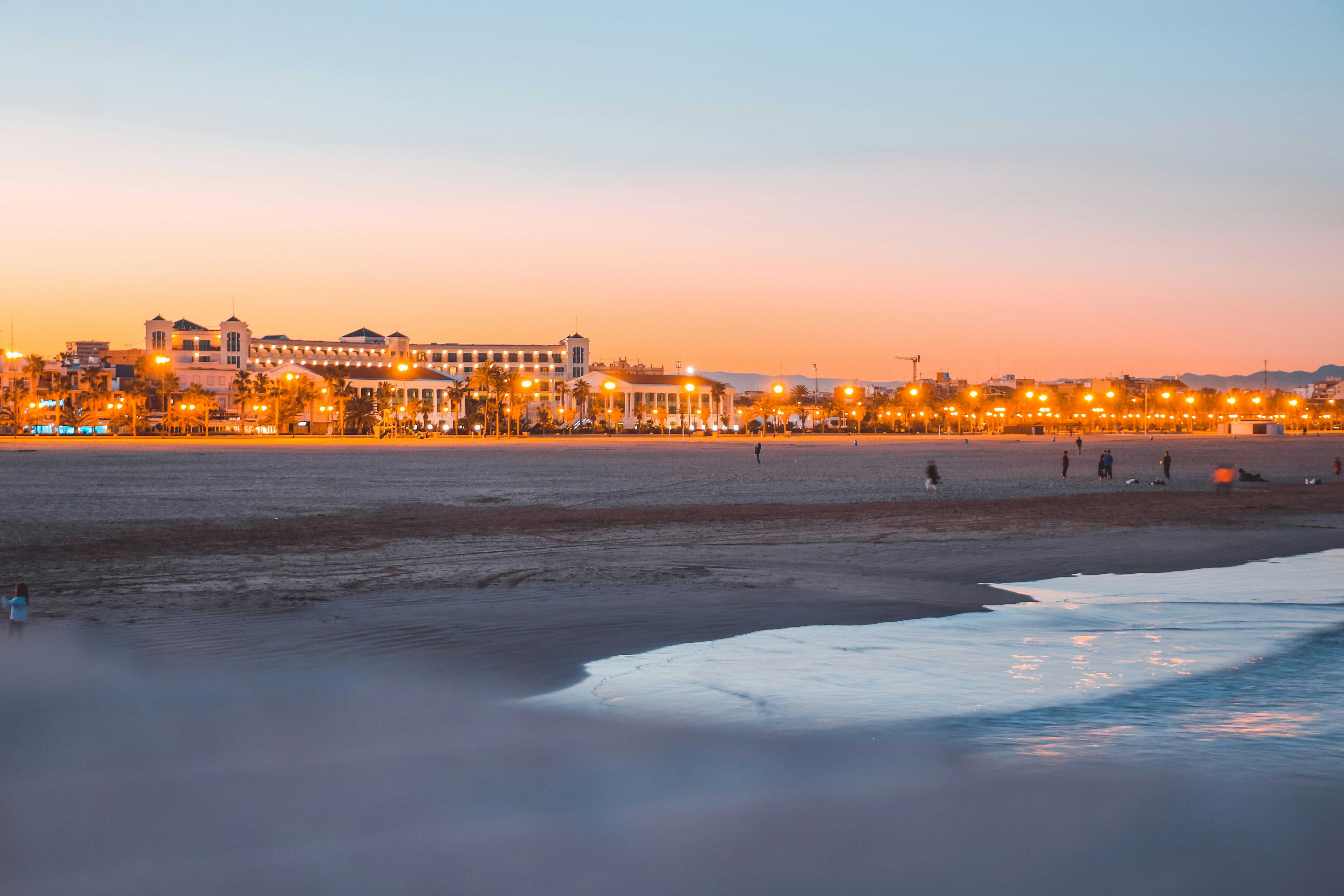 Illuminated beachfront with a hotel and gentle waves reflecting the twilight colors. Soft sand and distant figures create a serene evening atmosphere.