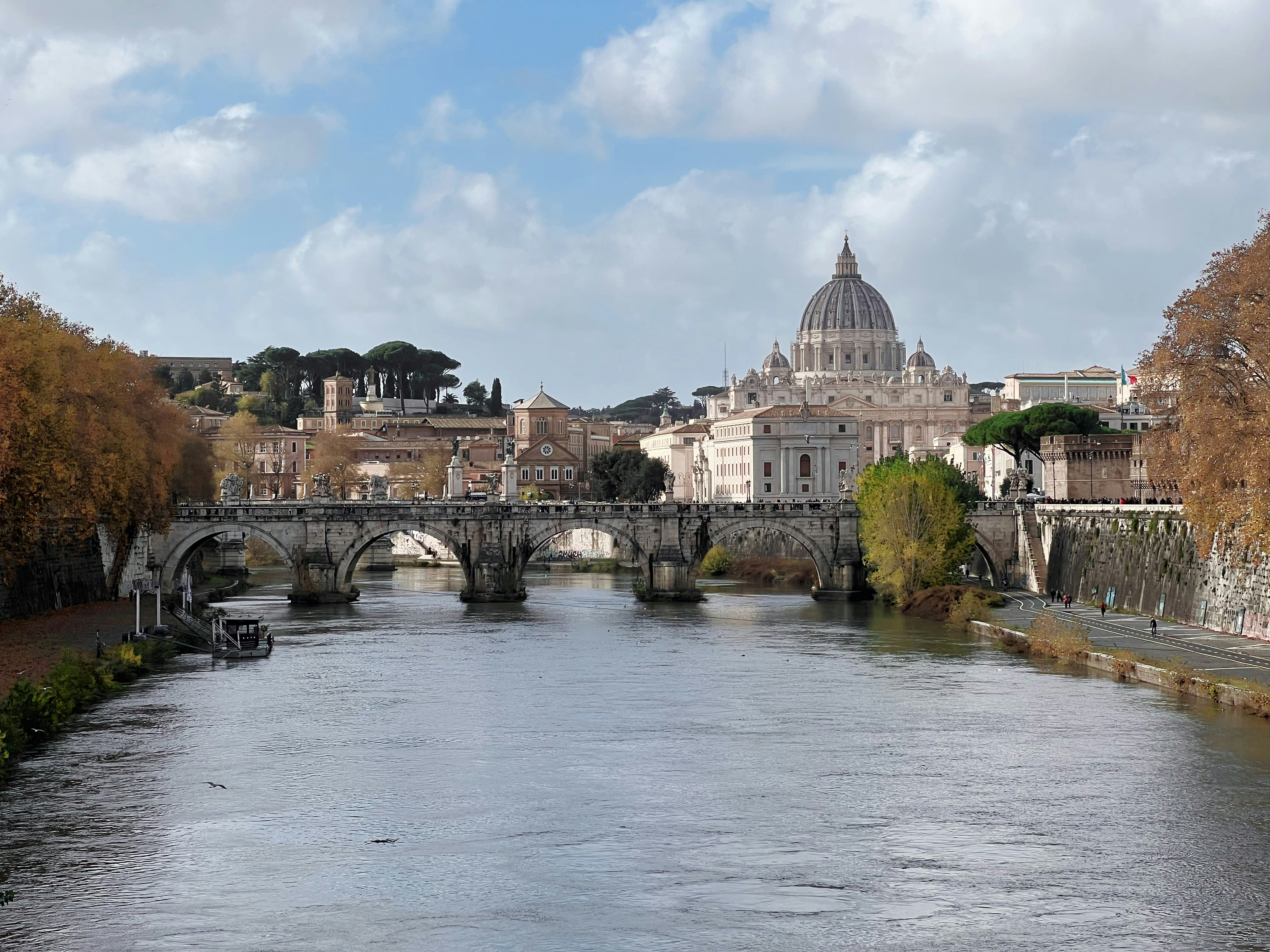 Historic bridge crossing the Tiber River with St. Peter's Basilica in the background, showcasing autumn foliage along the banks.