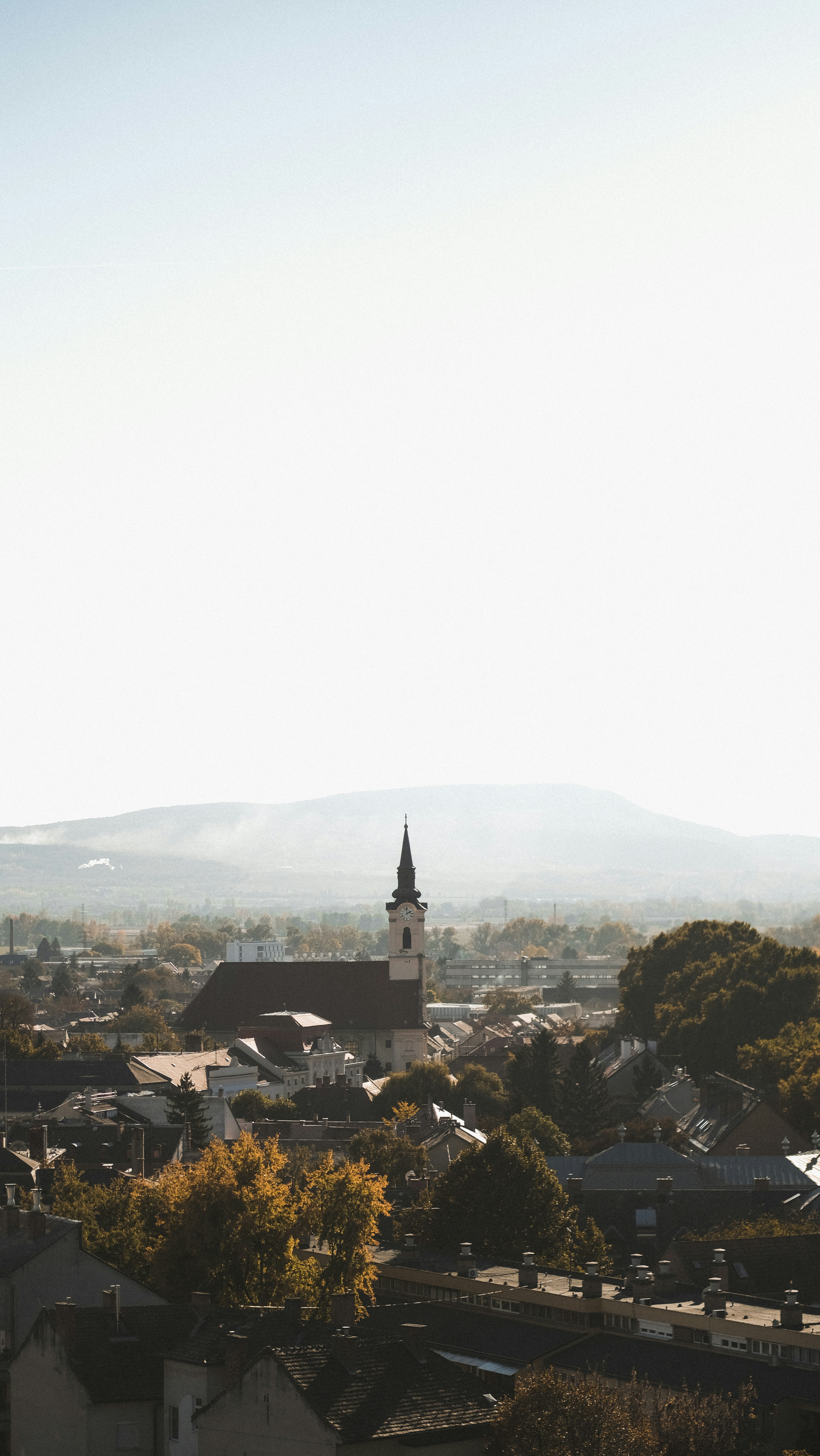 Historic church steeple rises above a tranquil village, framed by autumn foliage and distant mountains. Soft light bathes the scene in a gentle glow.