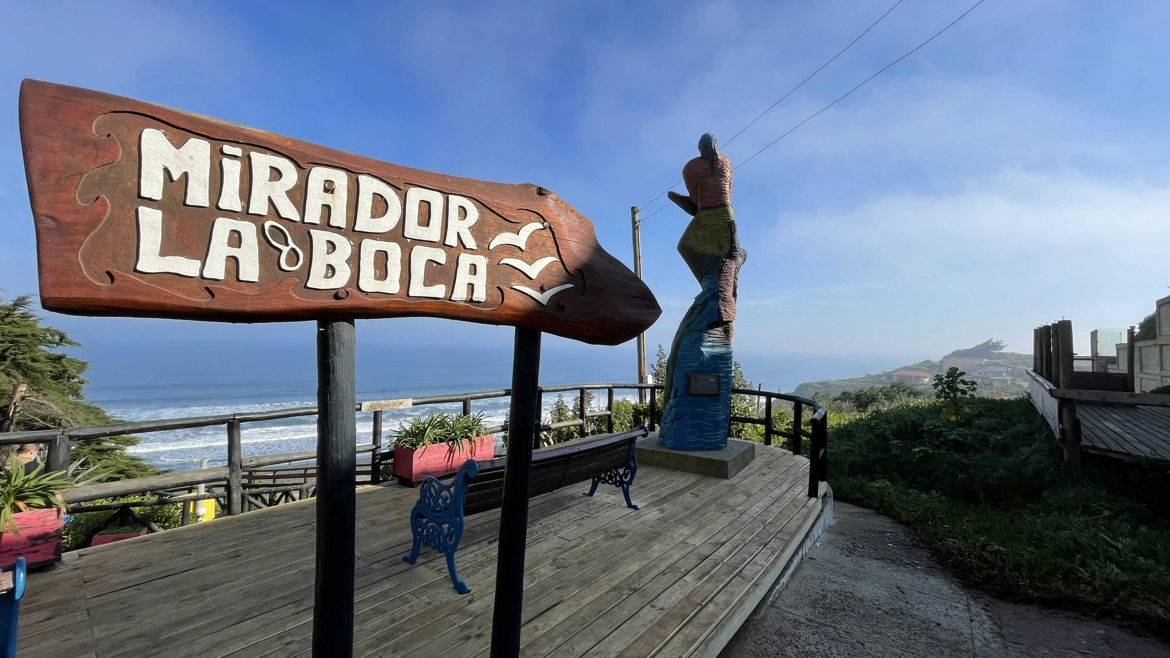 Wooden sign directing visitors to Mirador La Boca, accompanied by a colorful coastal sculpture, overlooking the ocean. 